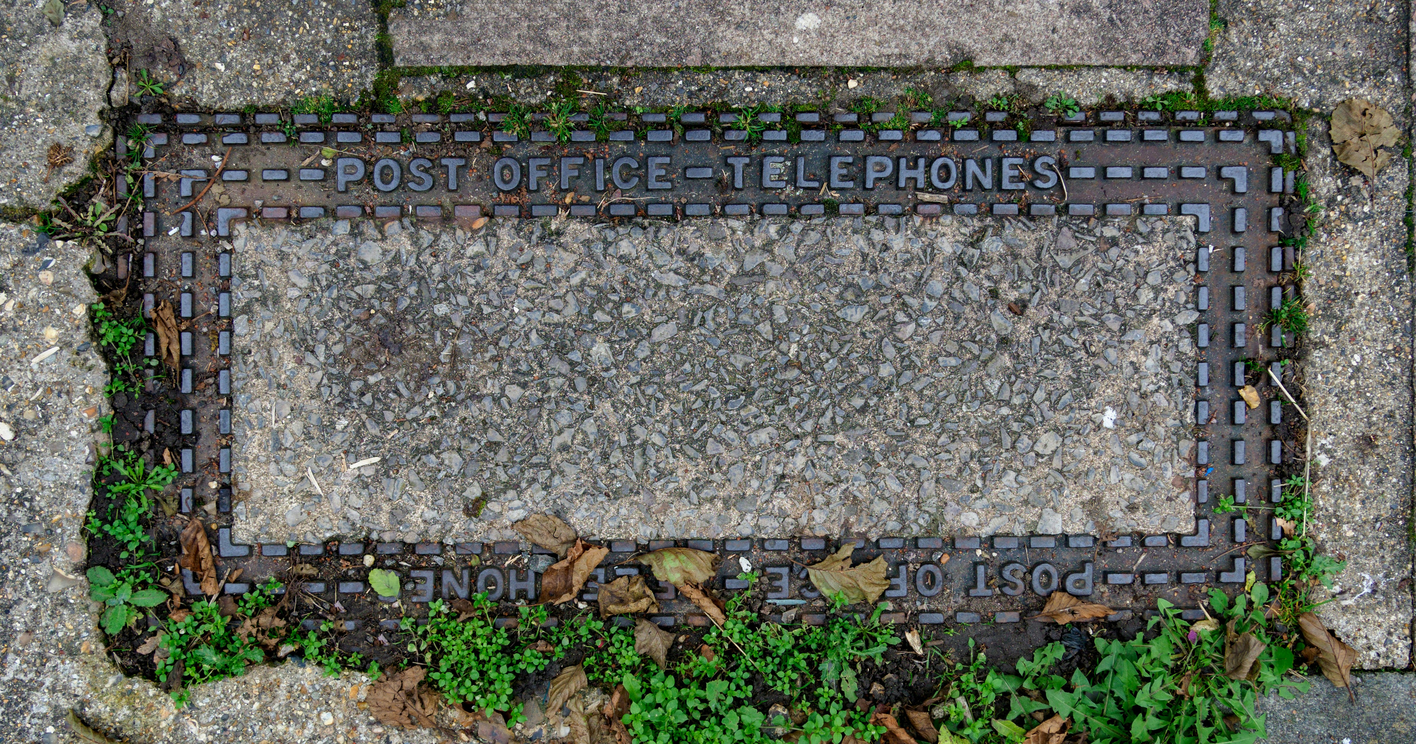 A weathered manhole cover inscribed with 'POST OFFICE TELEPHONES,' surrounded by patches of grass and fallen leaves.