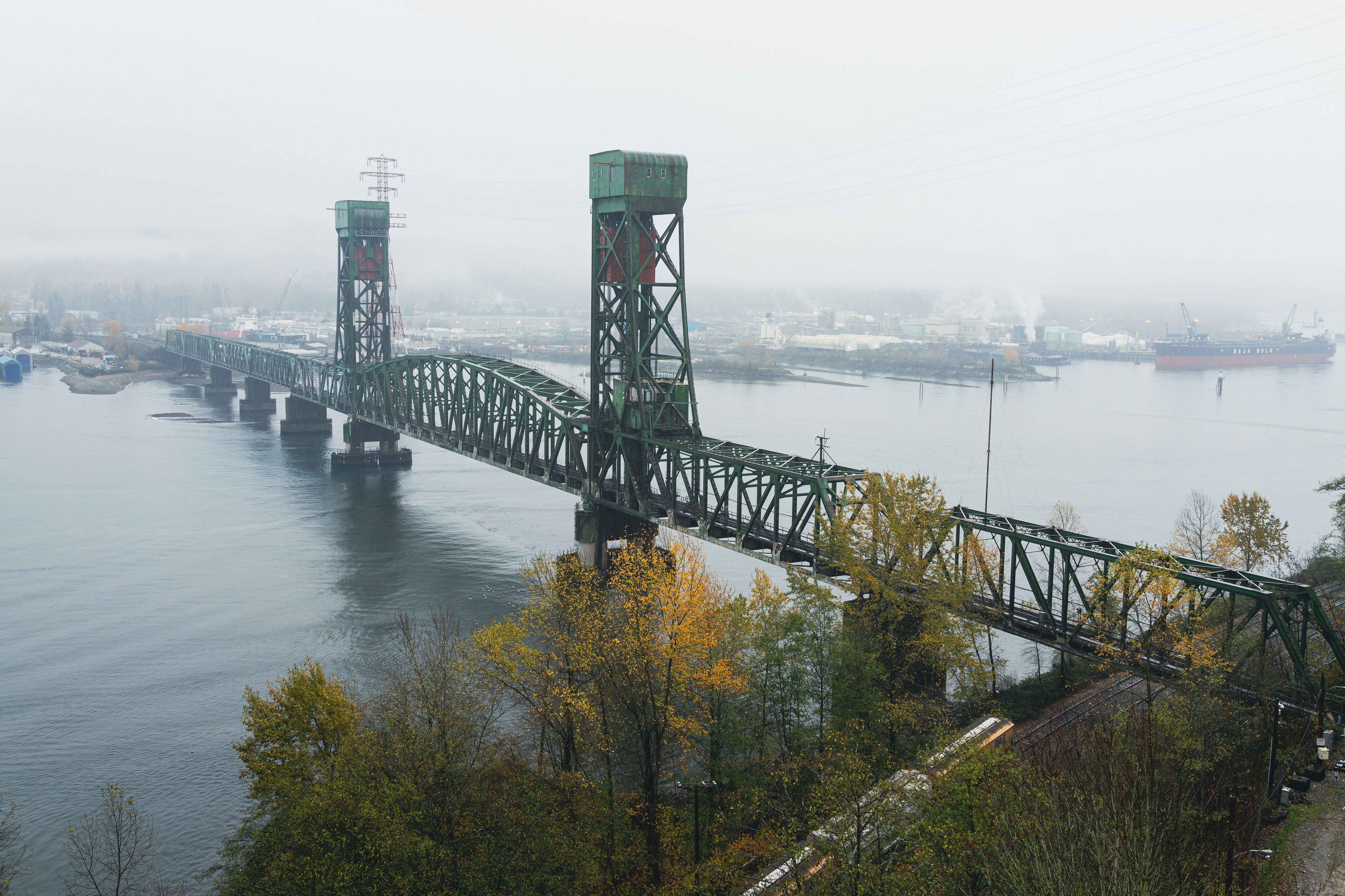 A steel bridge stands resilient amidst a foggy backdrop, flanked by autumn foliage and a tranquil river. The scene conveys a sense of quiet solitude.