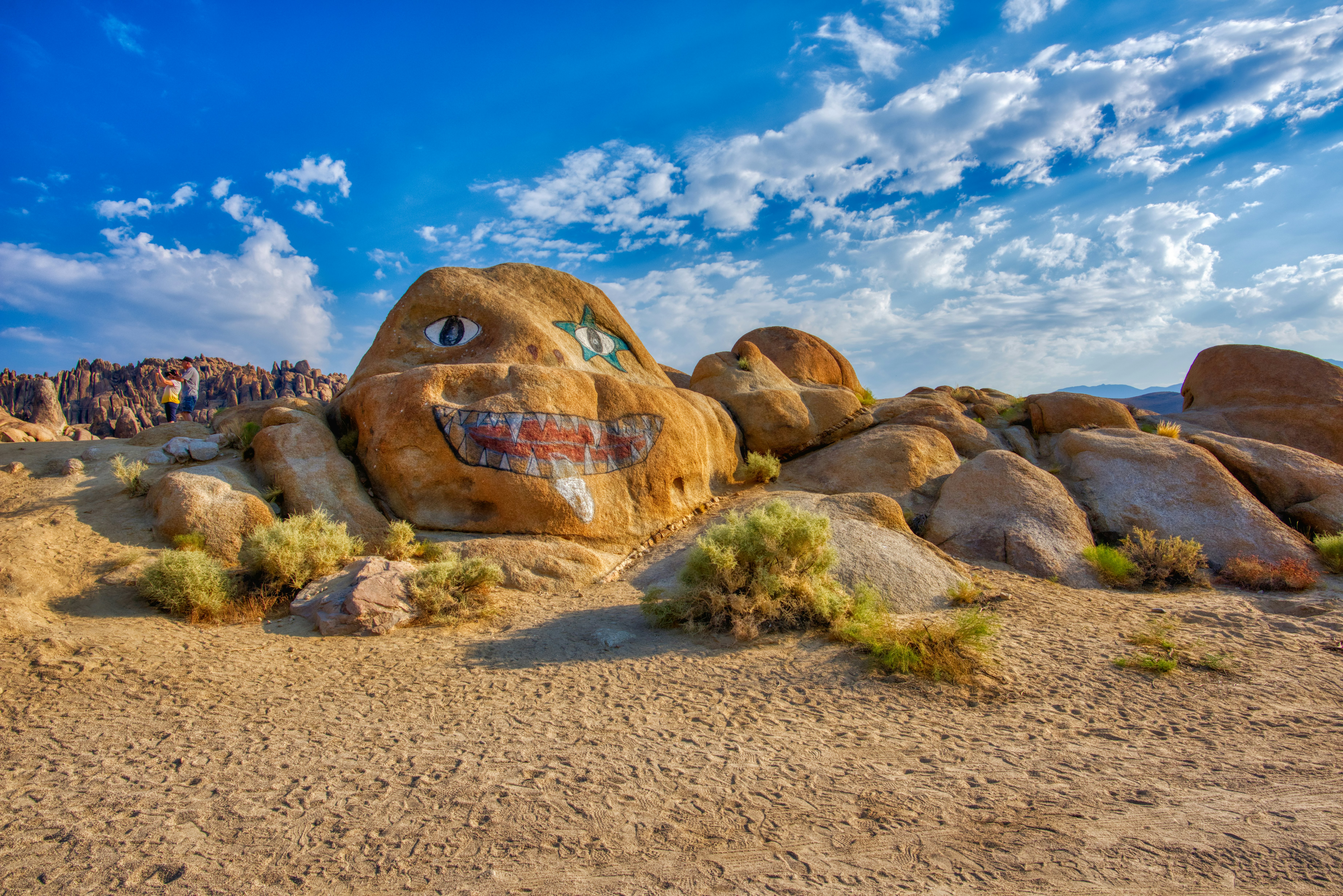 A large rock formation painted with a smiling face, surrounded by desert vegetation and dramatic skies. The playful artwork contrasts with the rugged landscape.