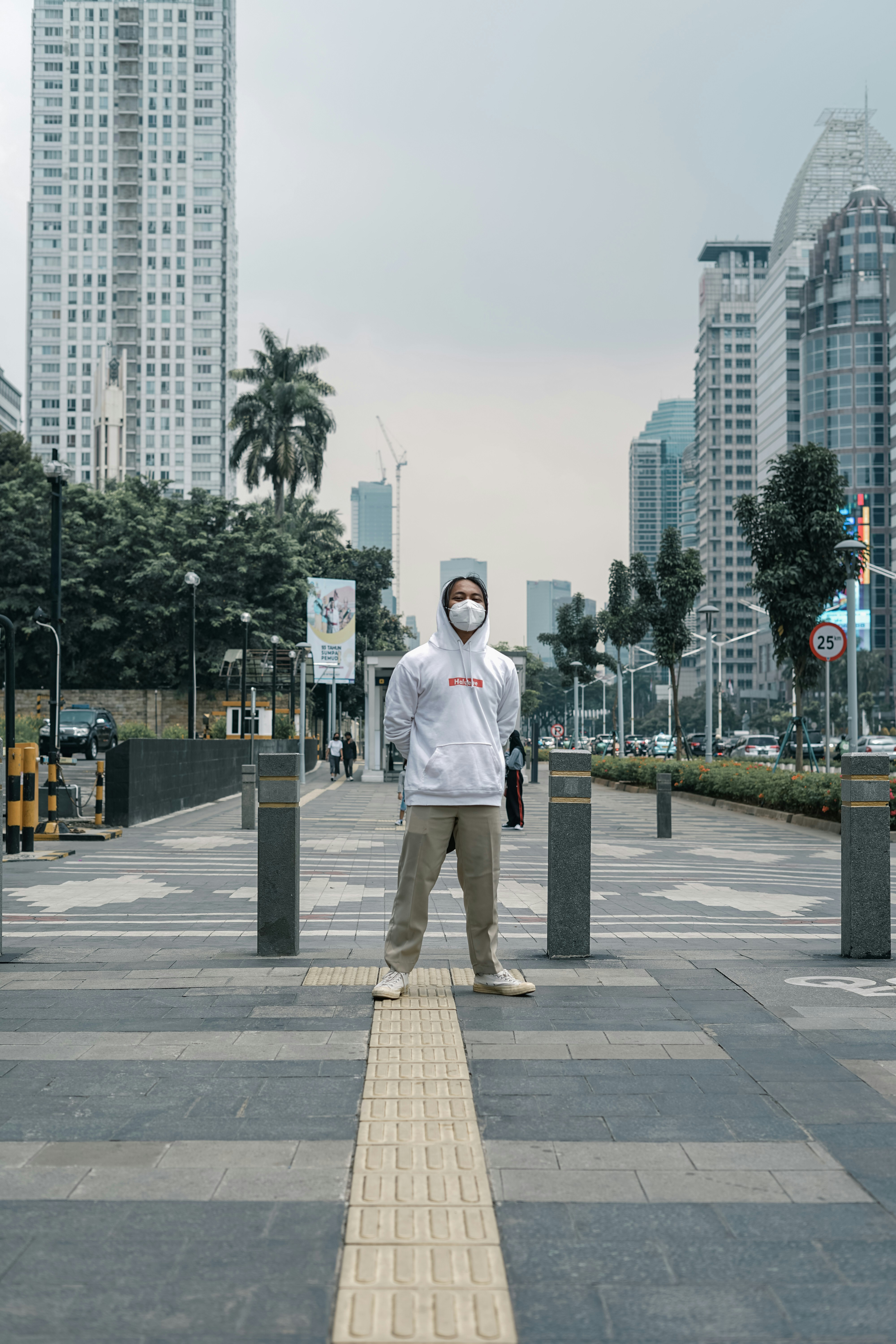 A person in a white hoodie stands on a city sidewalk, flanked by tall buildings and palm trees, evoking a sense of solitude amidst urban life.