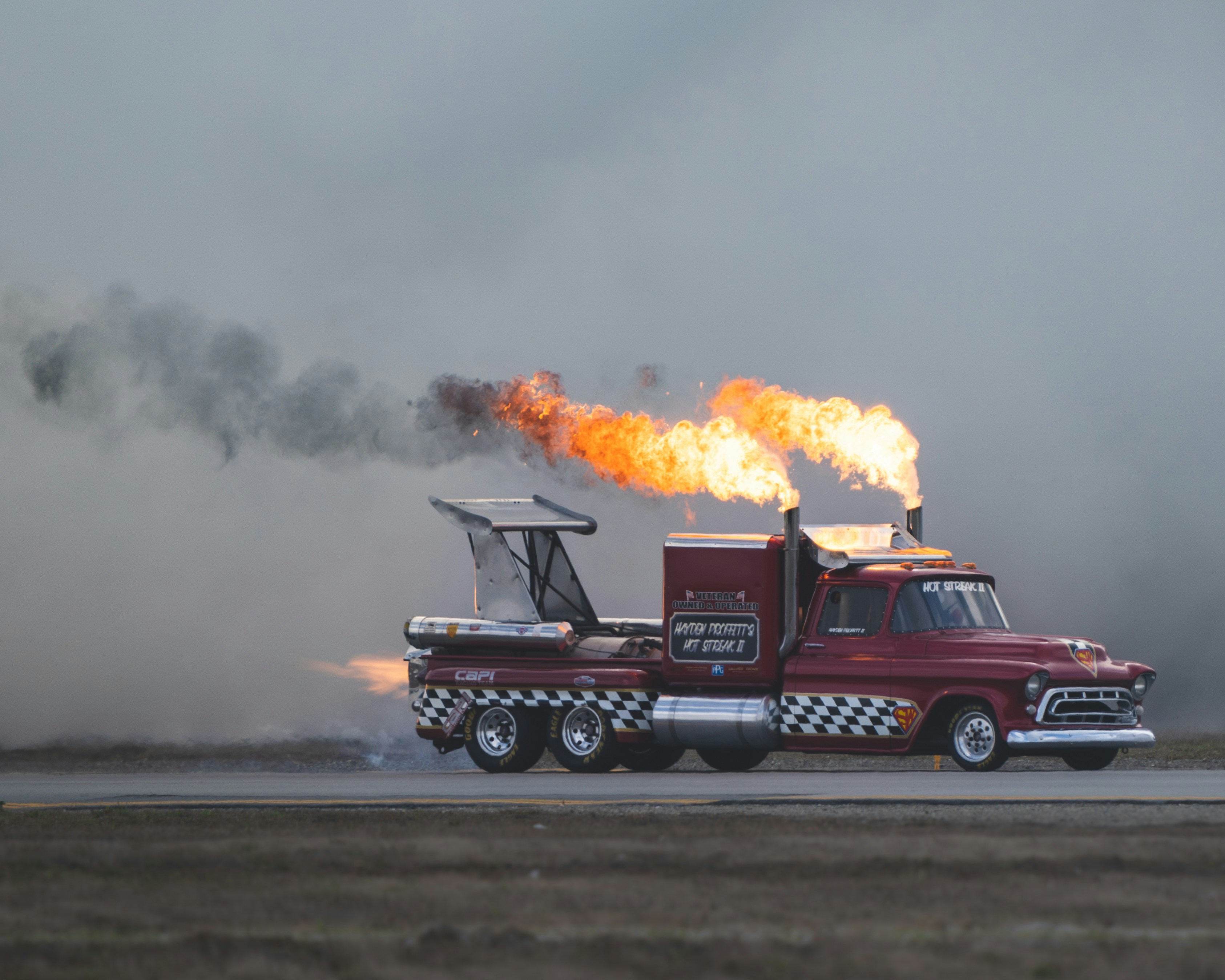 Vintage truck with flames erupting from its rear, set against a smoky backdrop during a performance event.