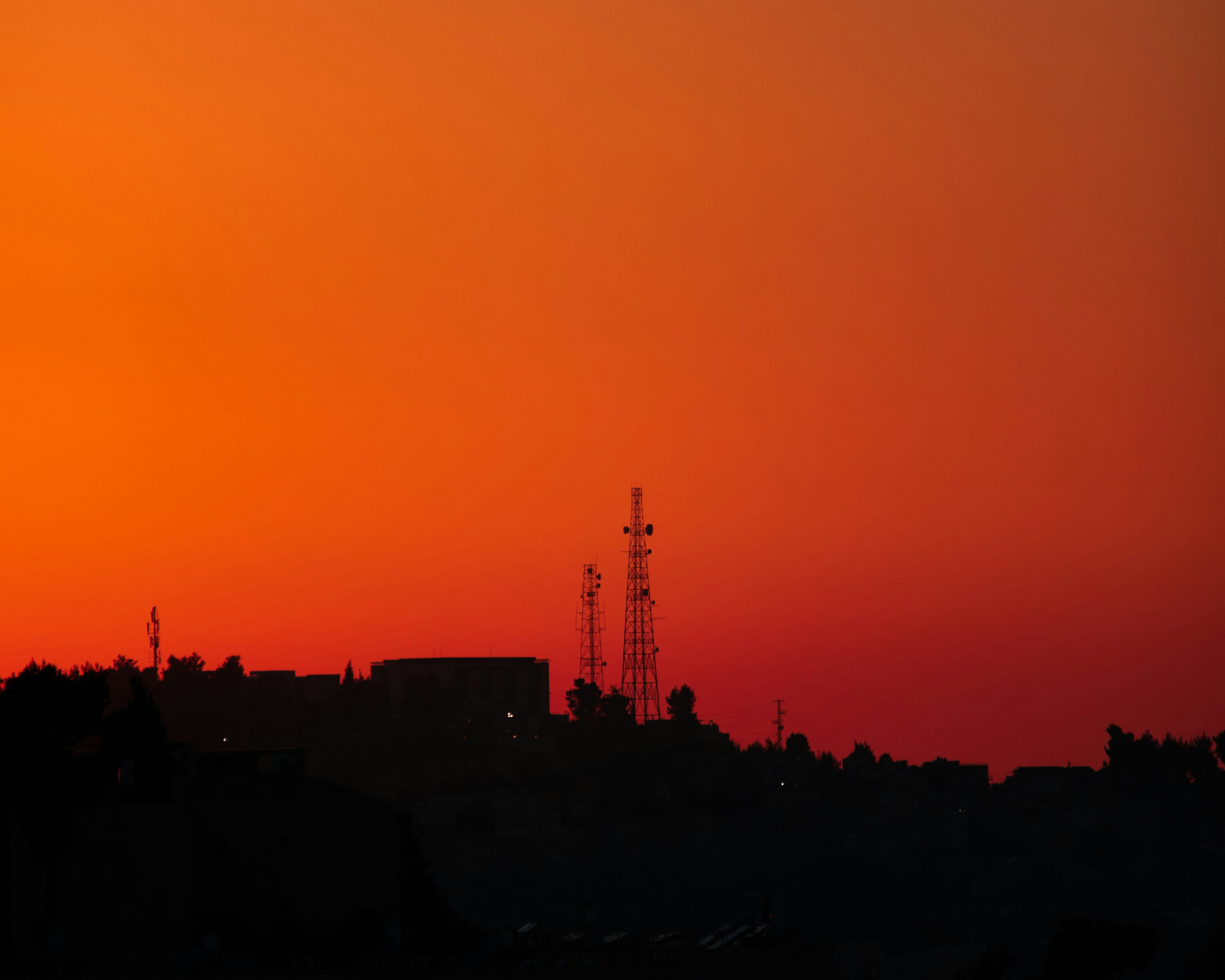 Silhouetted communication towers rise against a vibrant orange sunset, creating a stark contrast with the sky's gradient hues.