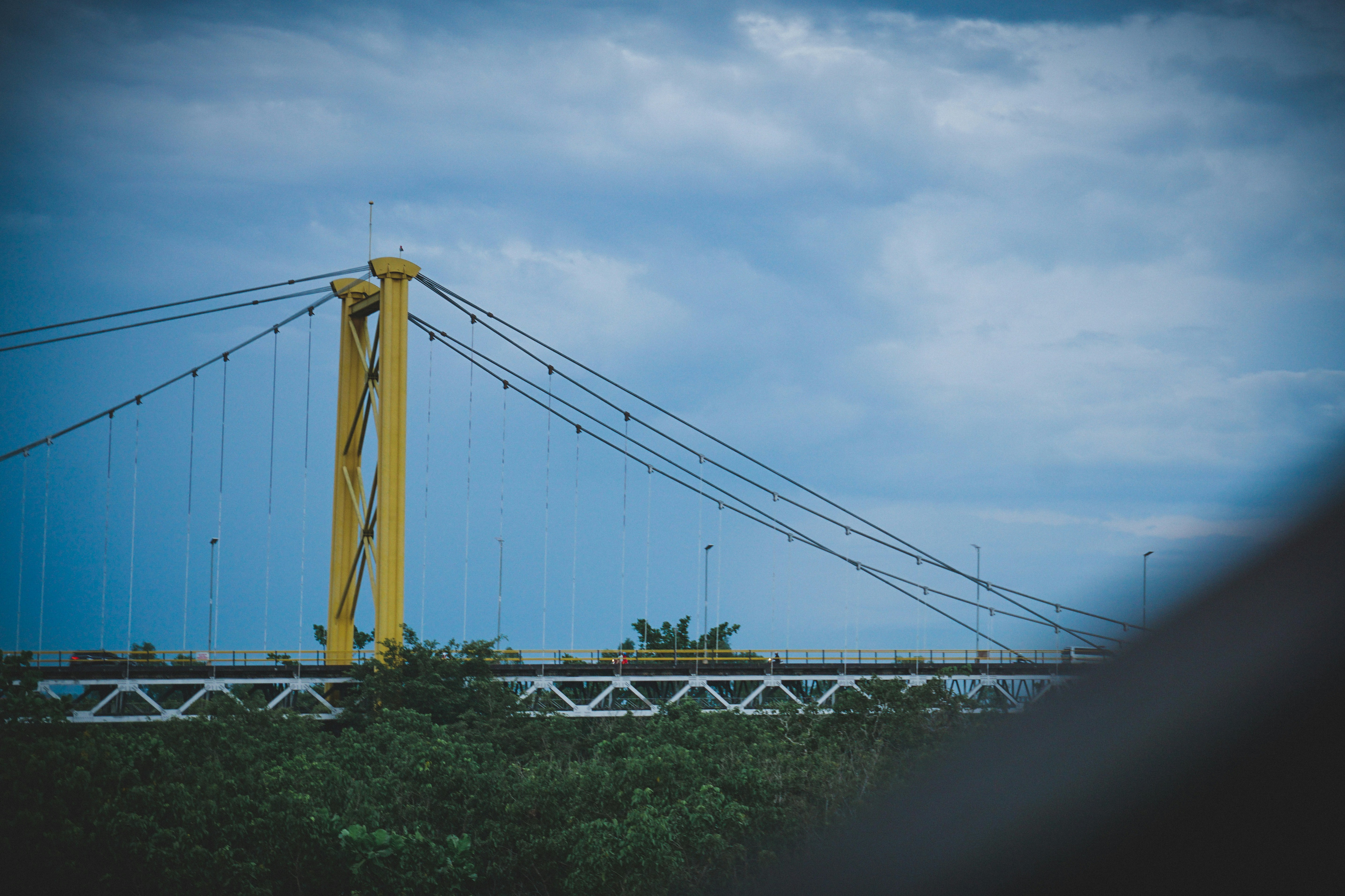 A vibrant yellow suspension bridge looms against a moody sky, framed by lush greenery below.
