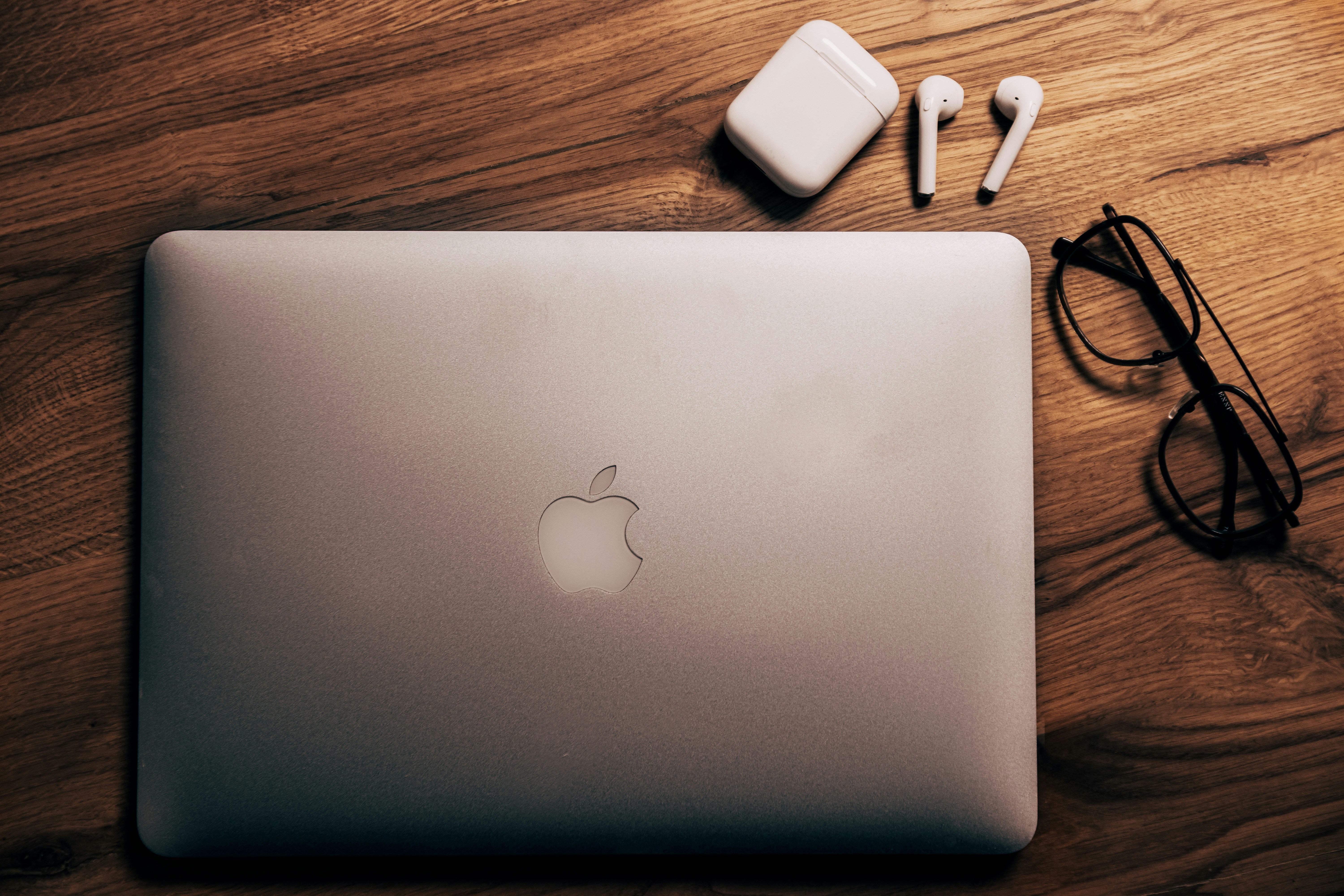 A sleek Apple laptop rests on a wooden surface, accompanied by wireless earbuds and a pair of glasses, embodying a modern workspace aesthetic.