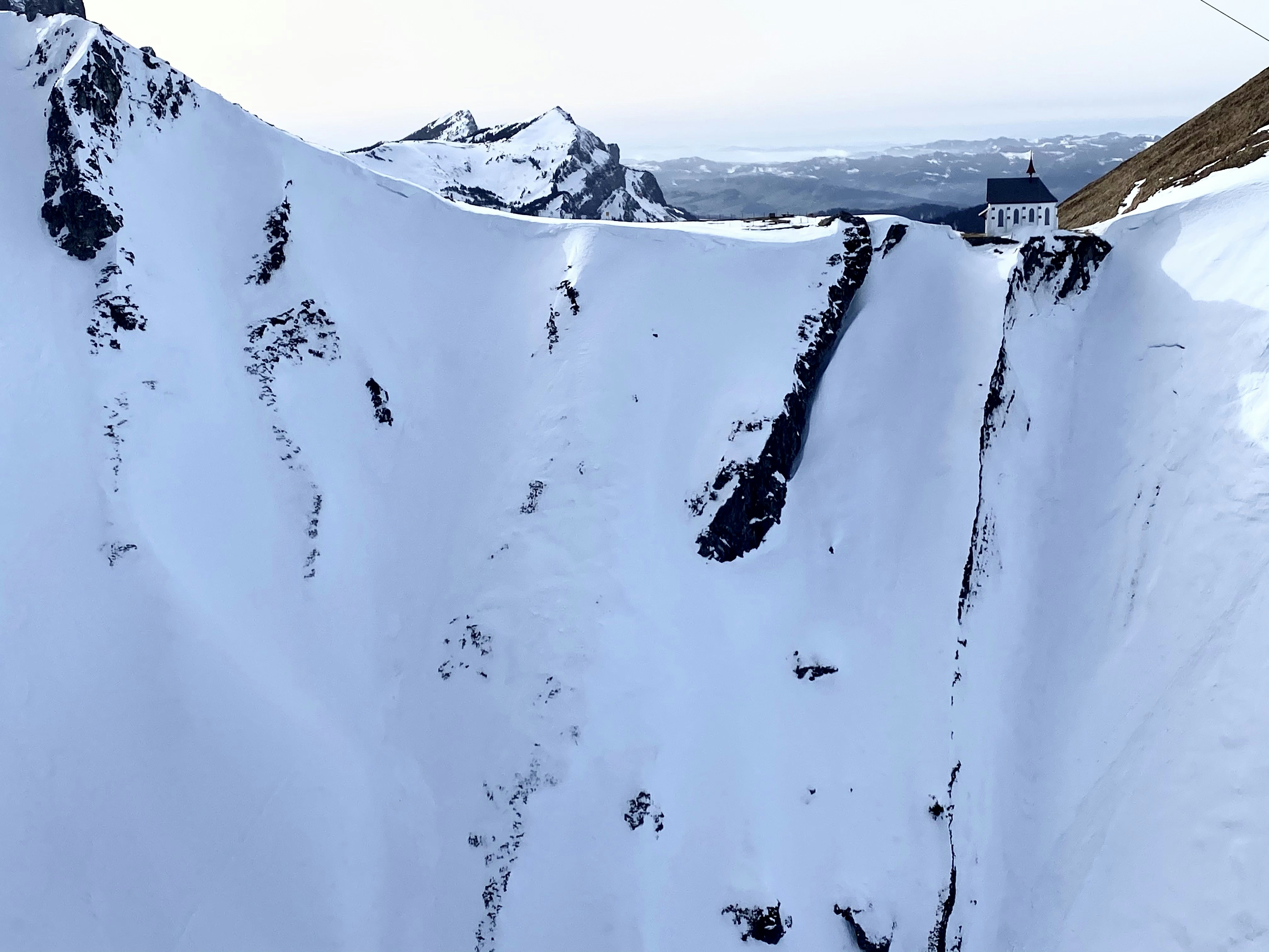 A solitary mountain cabin stands on a snowy cliff, overlooking a vast, rugged landscape. The stark contrast between the white snow and dark rock formations creates a dramatic scene.