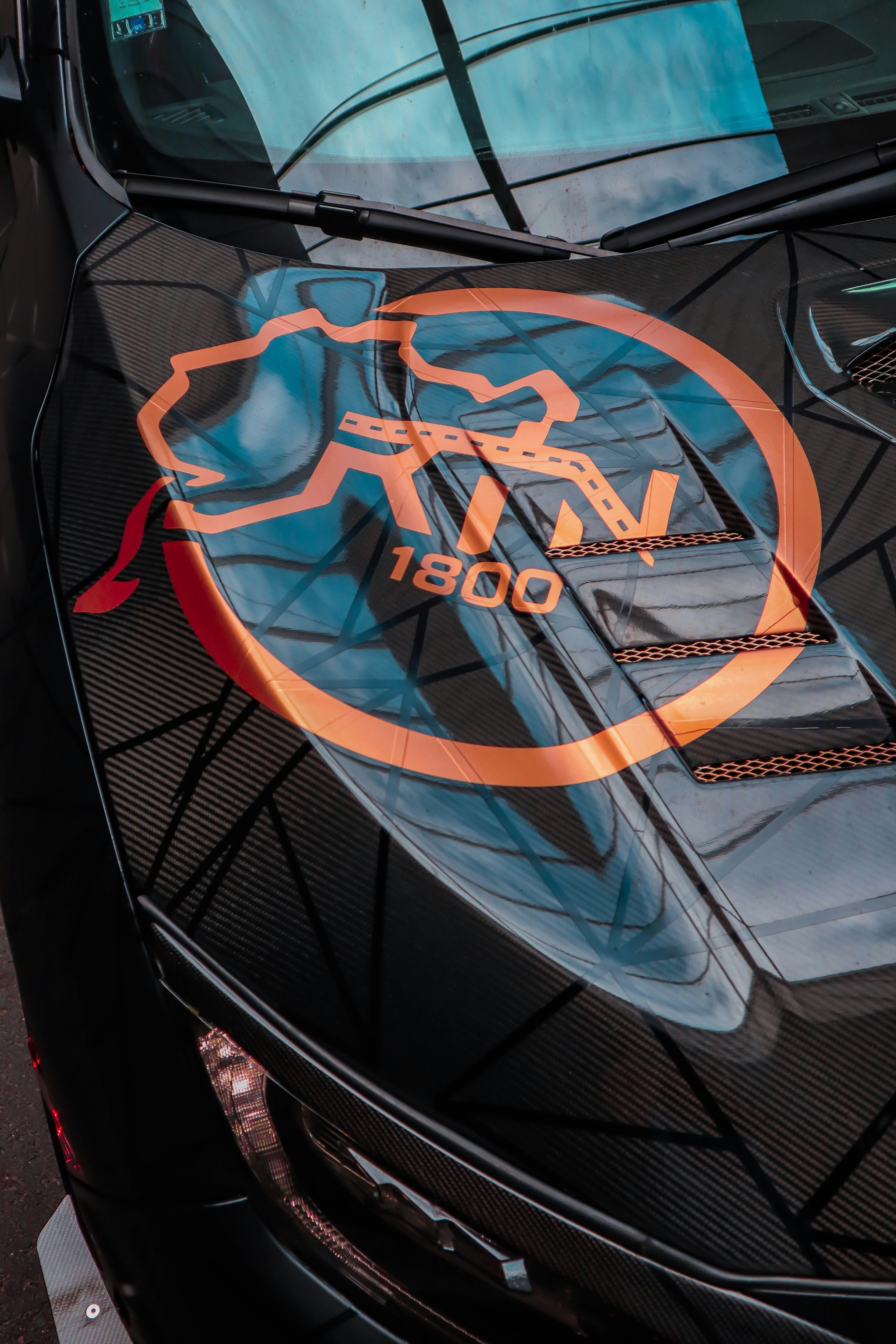 Orange Circular Emblem on Glossy Carbon-Fiber Hood Amid Sky Reflections