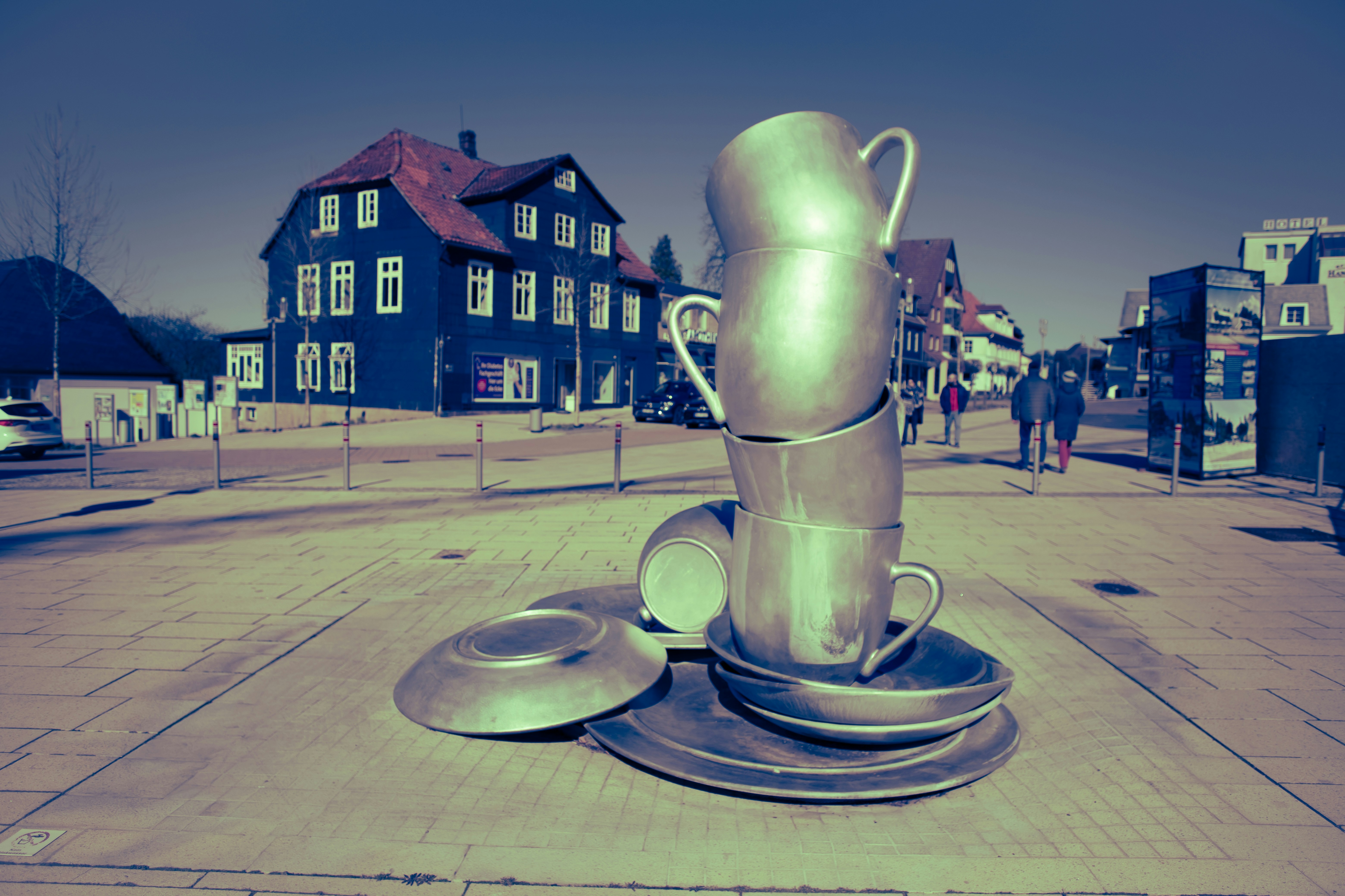 A whimsical sculpture of stacked metallic cups and saucers in an urban plaza, surrounded by buildings and pedestrians.