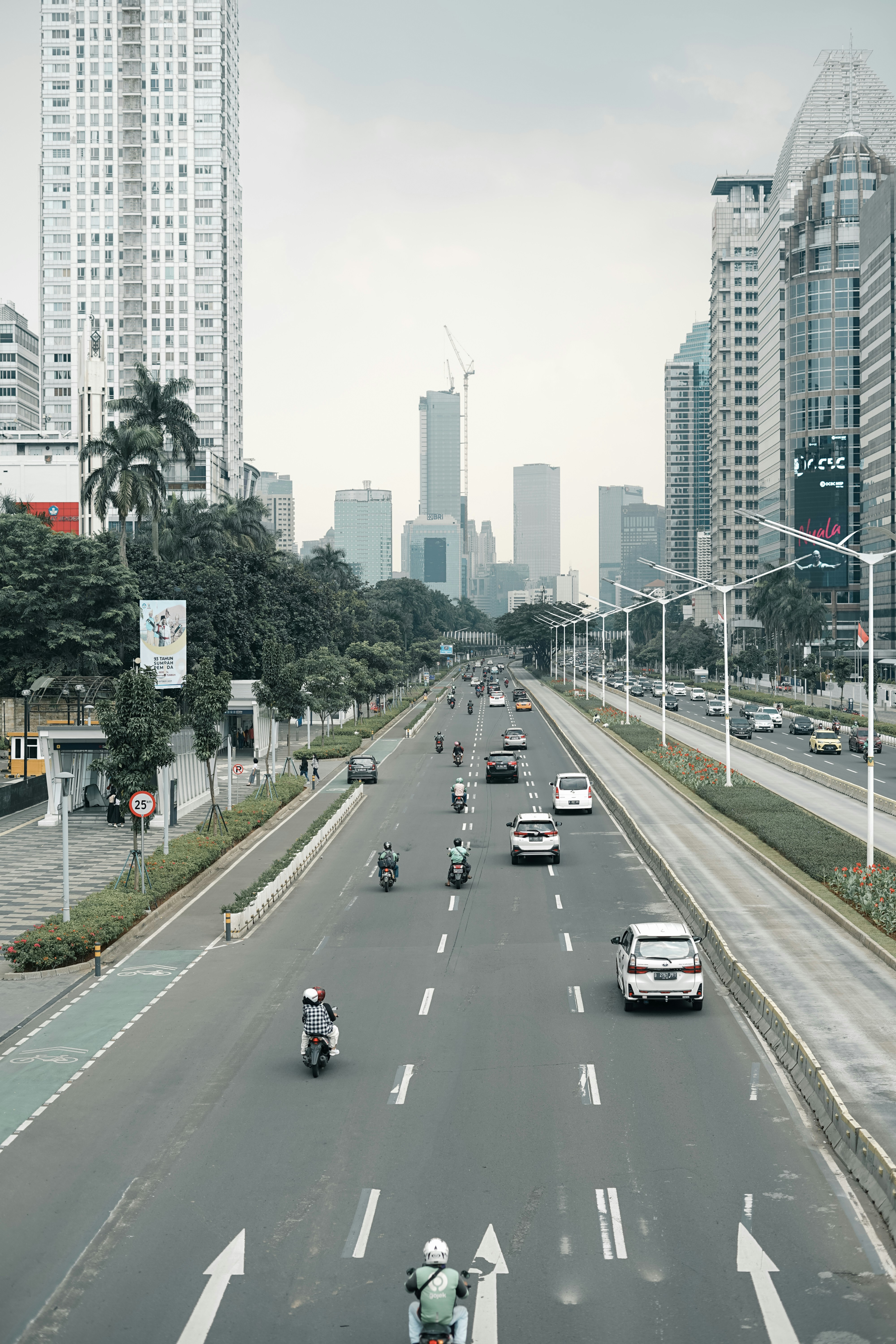 Busy urban road lined with modern skyscrapers, featuring a mix of cars and motorcycles navigating through the cityscape.