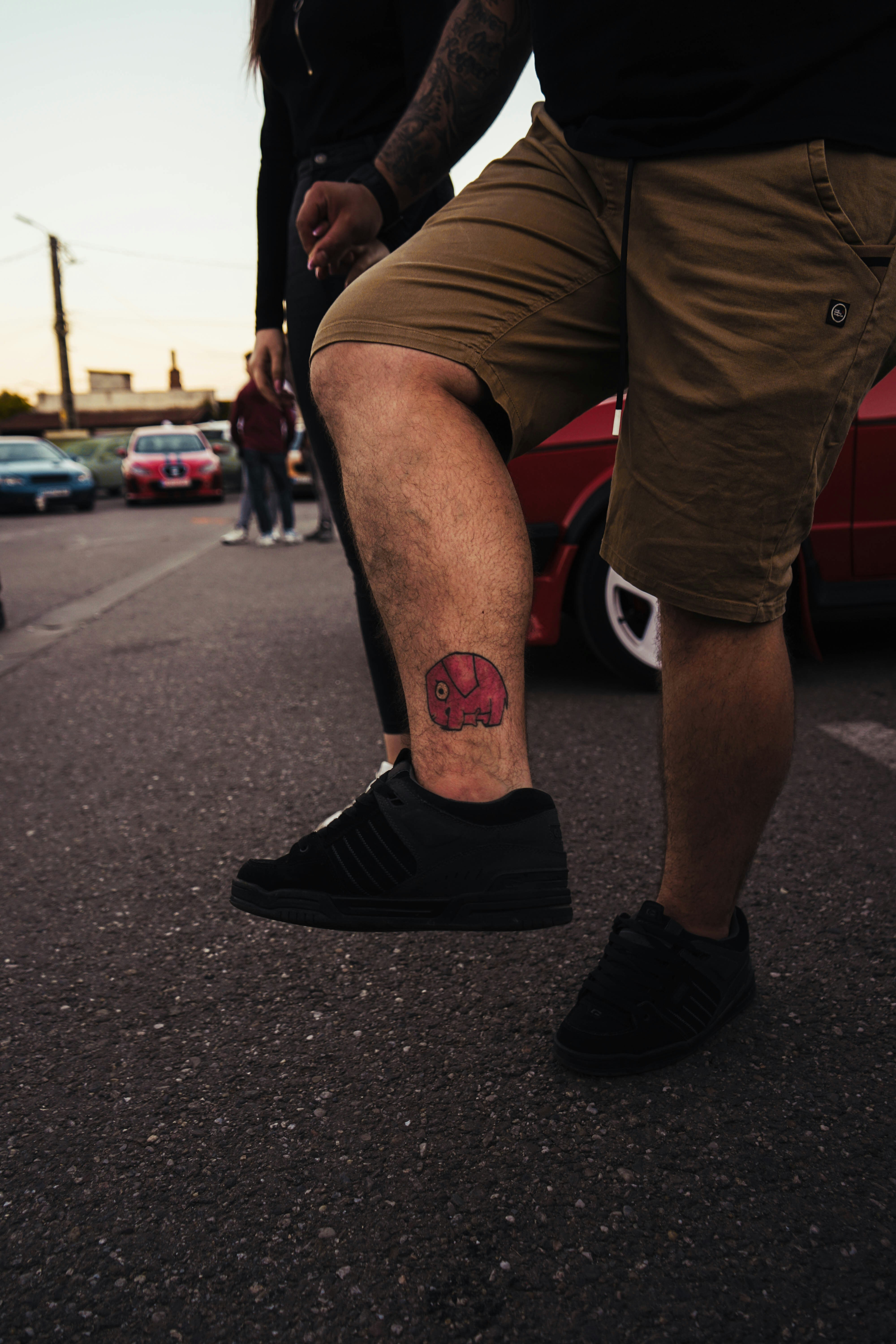 A close-up of a man's leg showcasing a colorful elephant tattoo, set against a backdrop of parked cars in an urban environment.