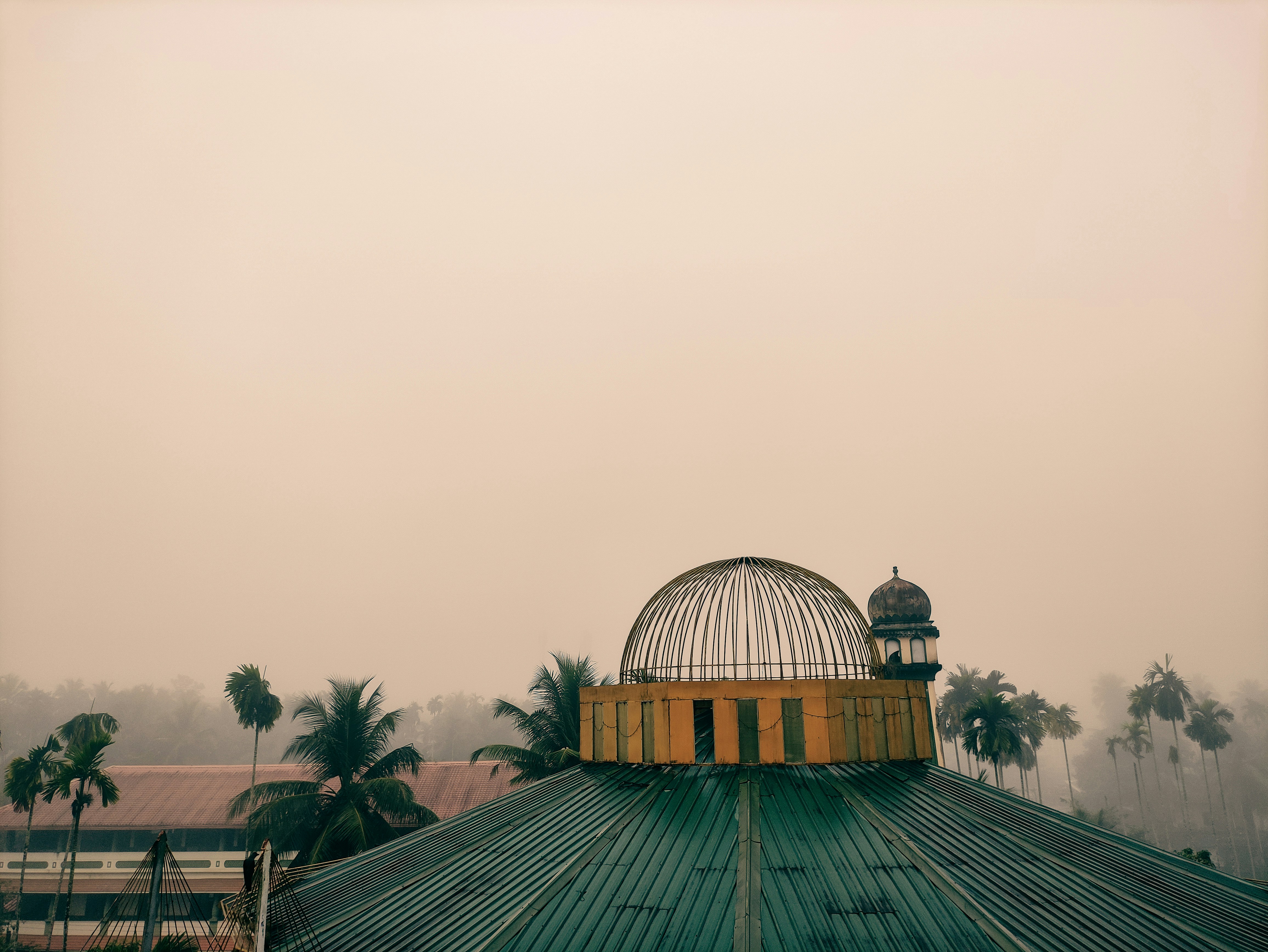 Photograph of a domed structure atop a corrugated rooftop with palm trees in hazy light.