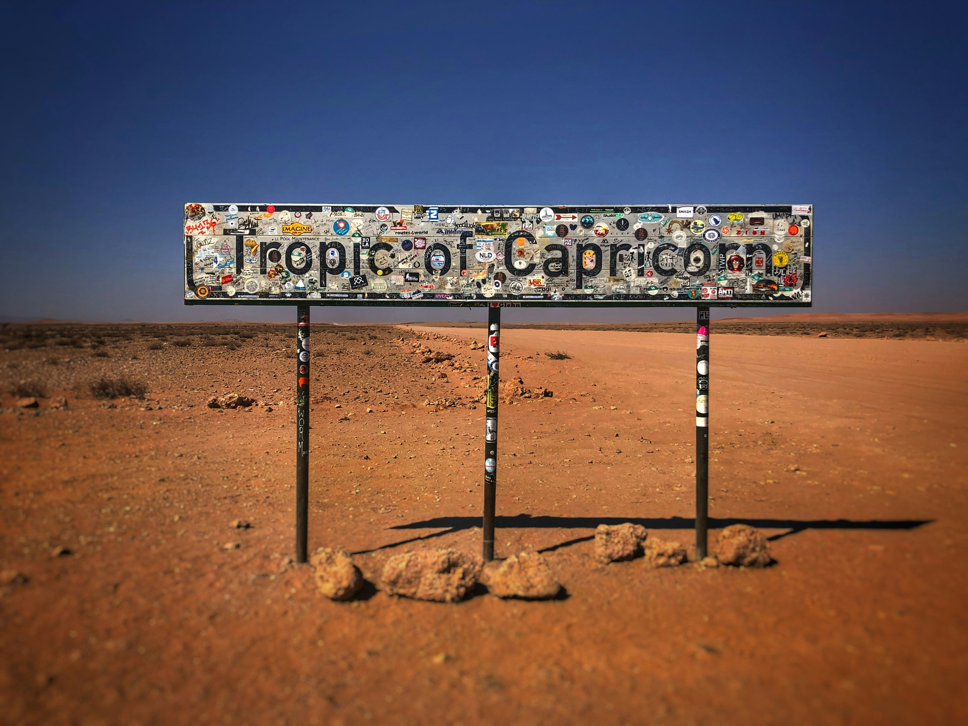 A weathered sign marking the Tropic of Capricorn, adorned with a variety of colorful stickers, set against a vast desert landscape. 