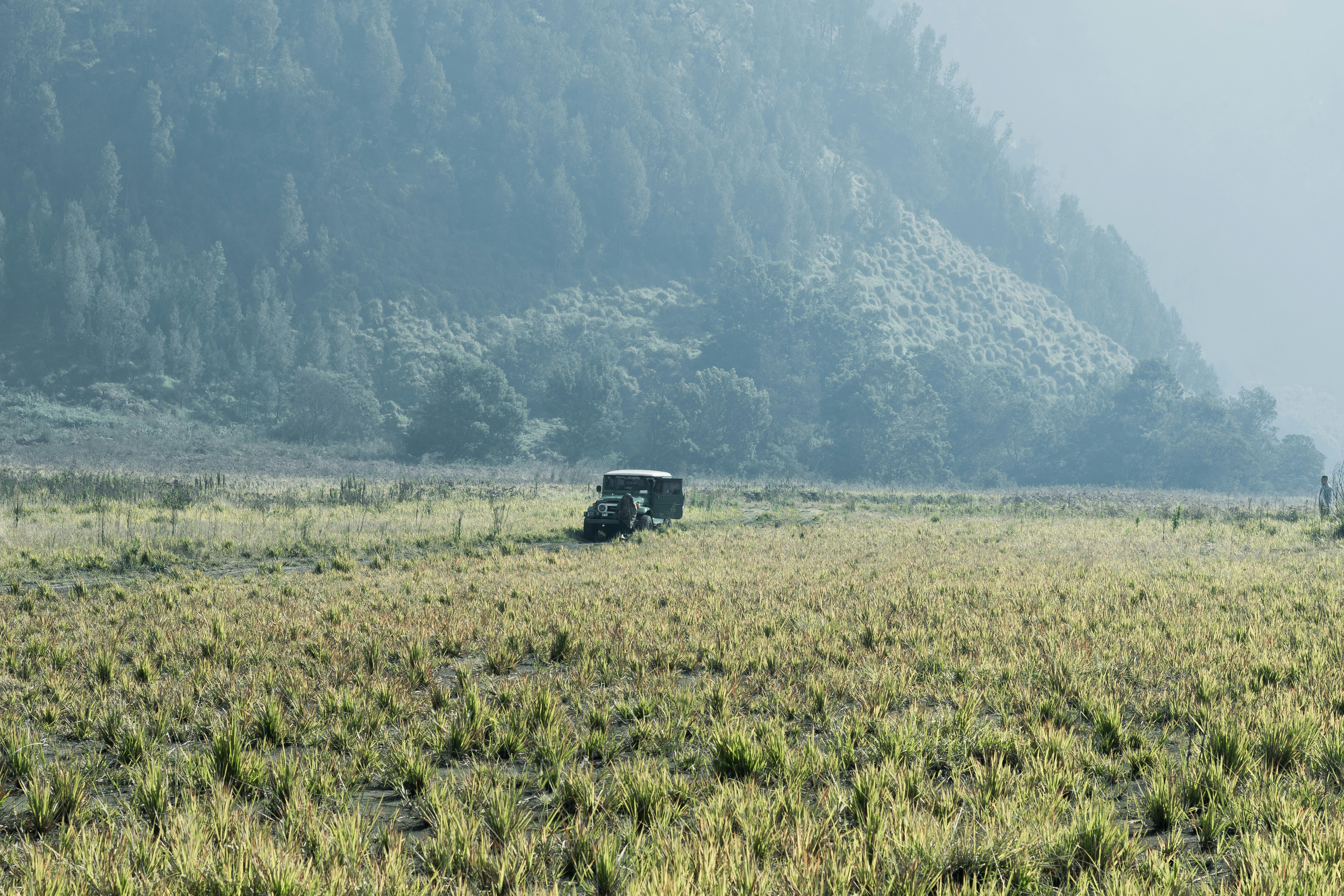 A vintage vehicle traversing a misty expanse of grassland, framed by distant hills and trees. The atmosphere conveys a sense of solitude and exploration.