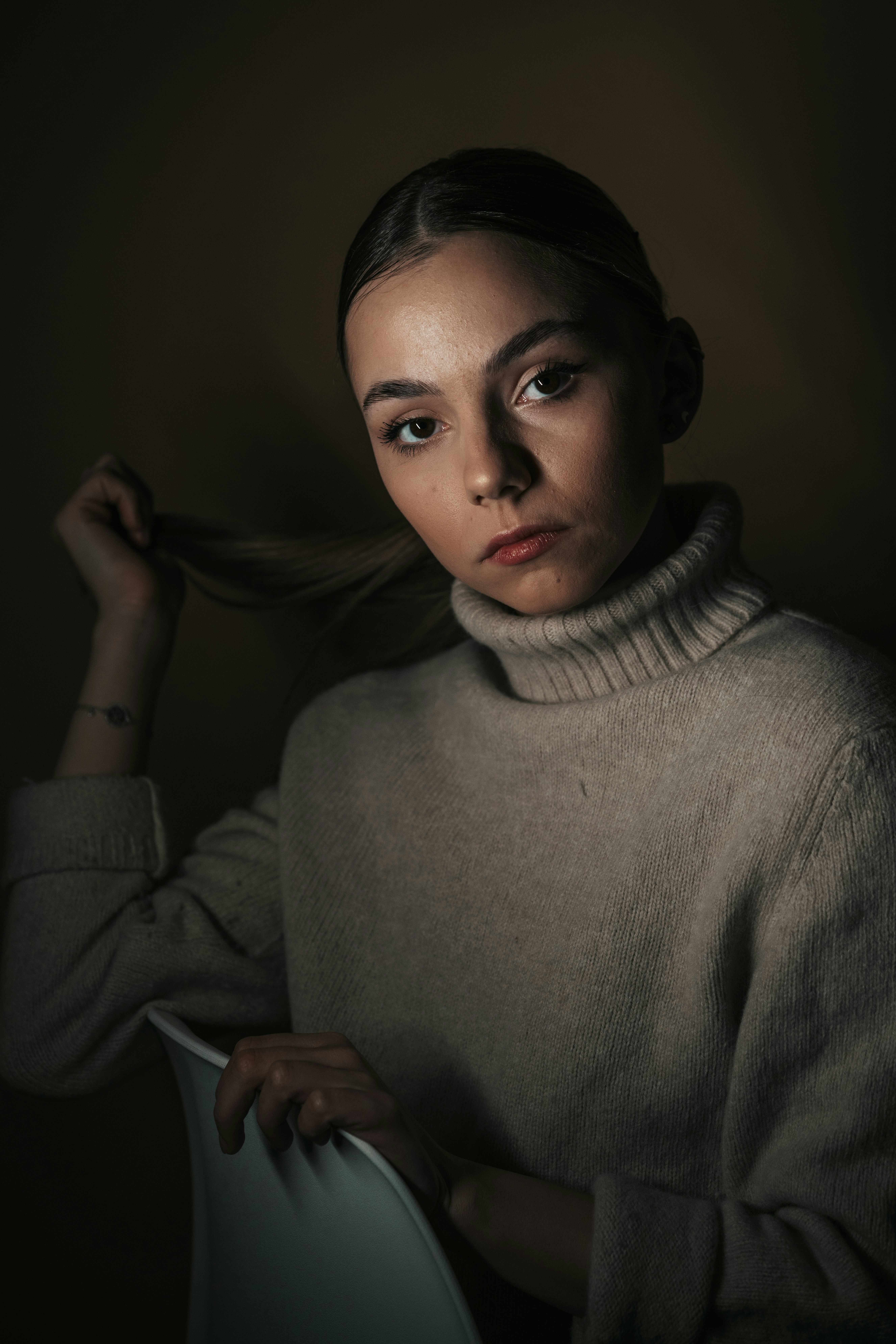 A contemplative young woman with long hair pulls at her ponytail while wearing a cozy turtleneck sweater against a warm backdrop.