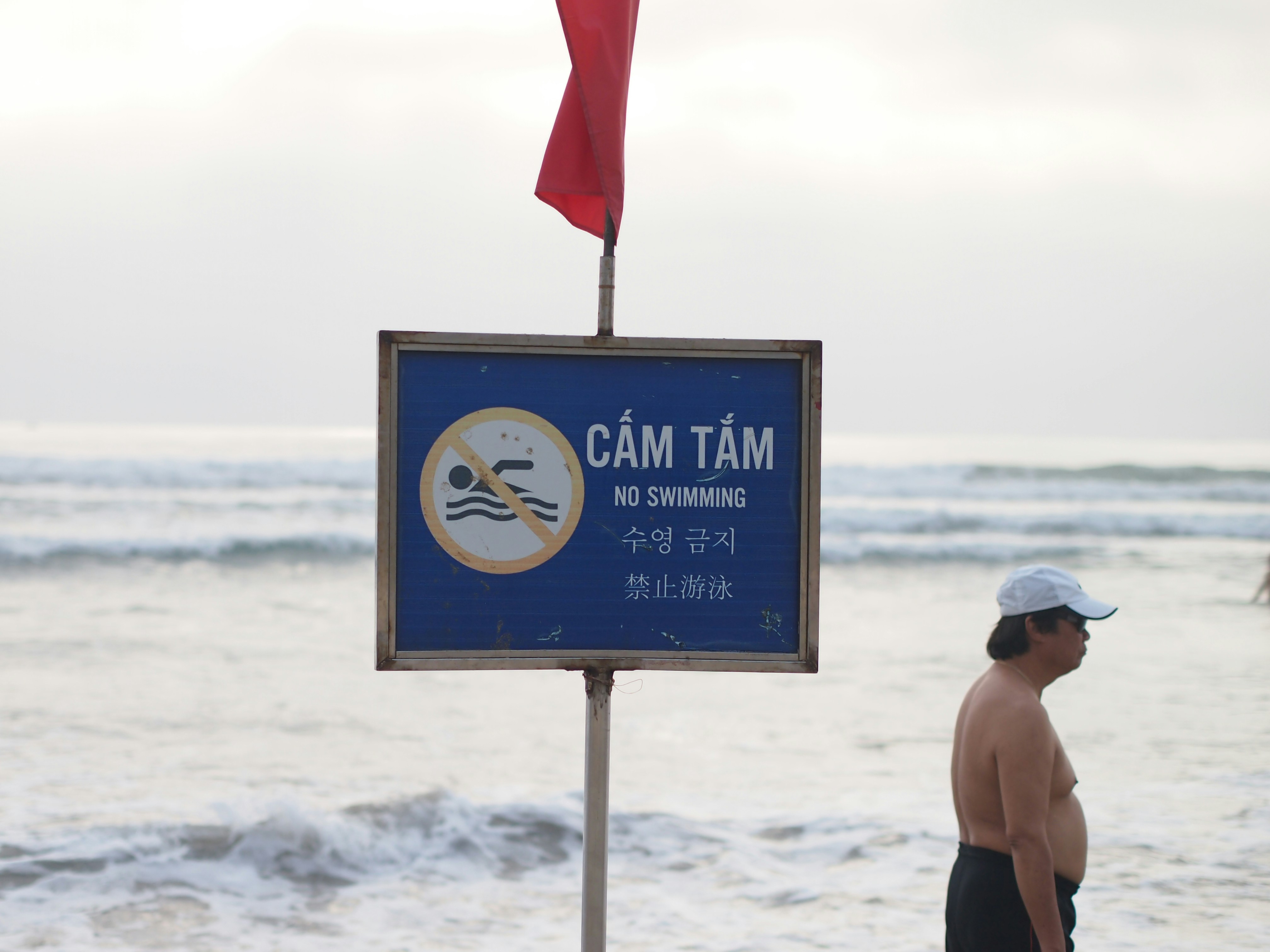 Photograph of a blue No Swimming sign on a beach with a shirtless man walking in the background.
