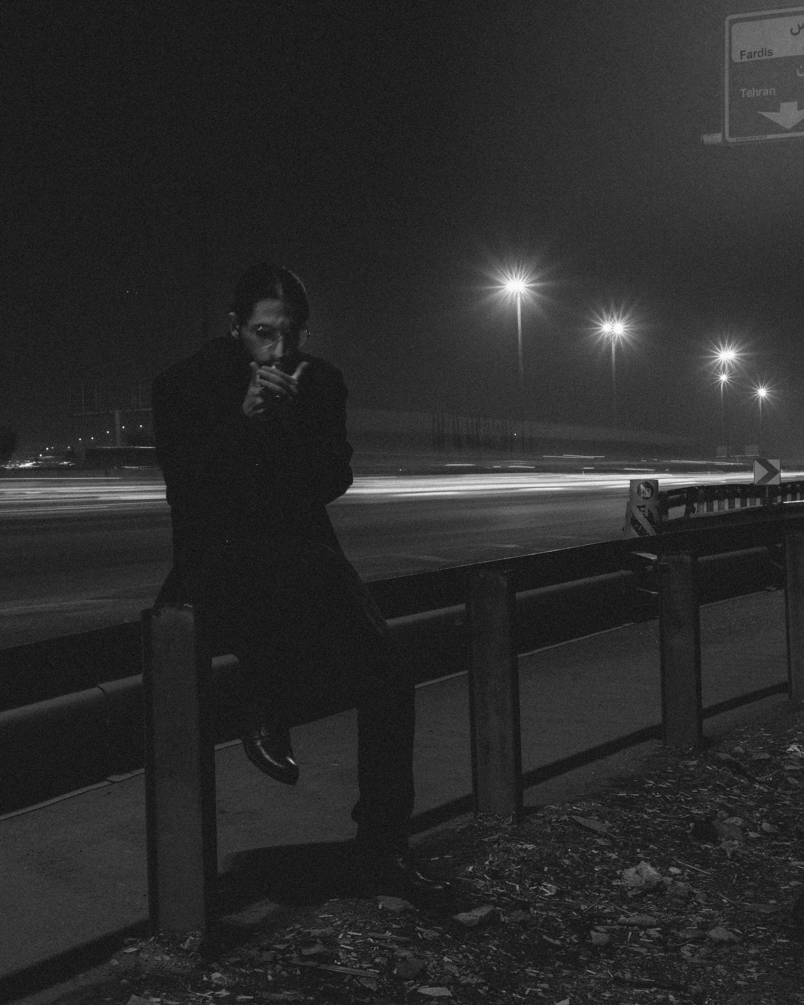 A figure in a black coat sits pensively on a railing at night, shrouded in shadows, with blurred lights of passing traffic illuminating the scene.