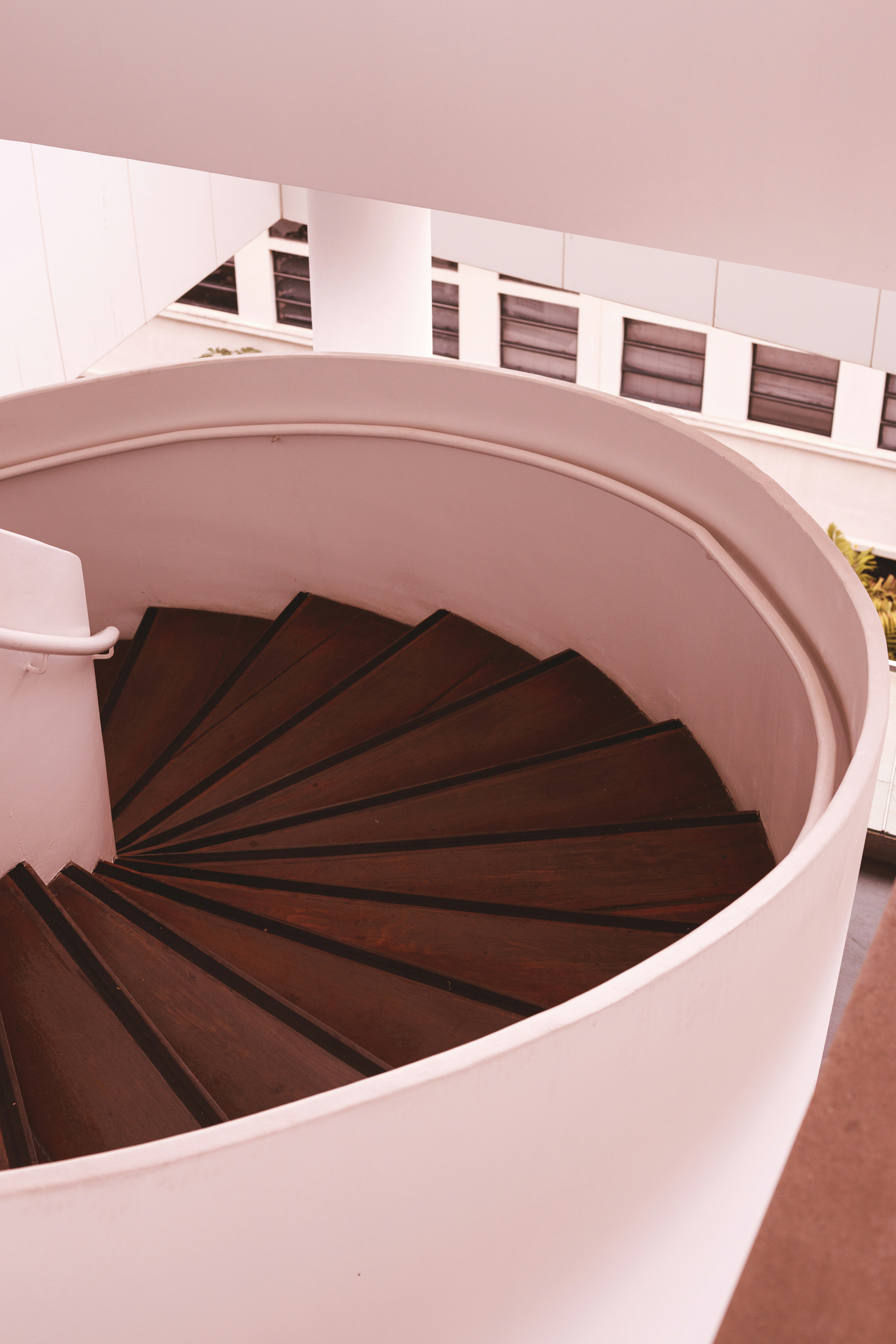 A circular wooden staircase spirals downward, framed by a smooth, white wall and soft natural light filtering through nearby windows.