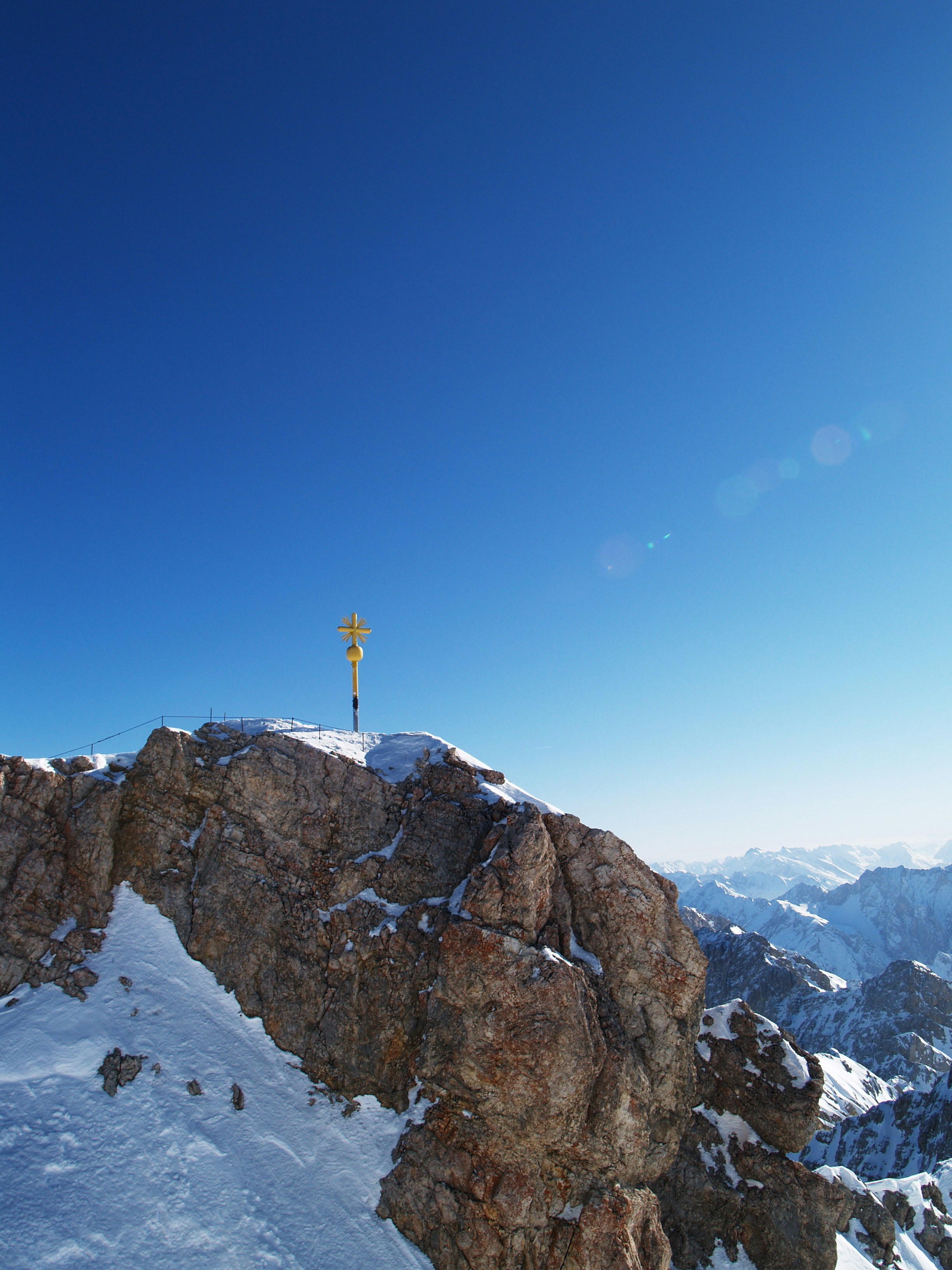 Snowy mountain peak with a solitary cross at the summit rises against a clear blue sky.