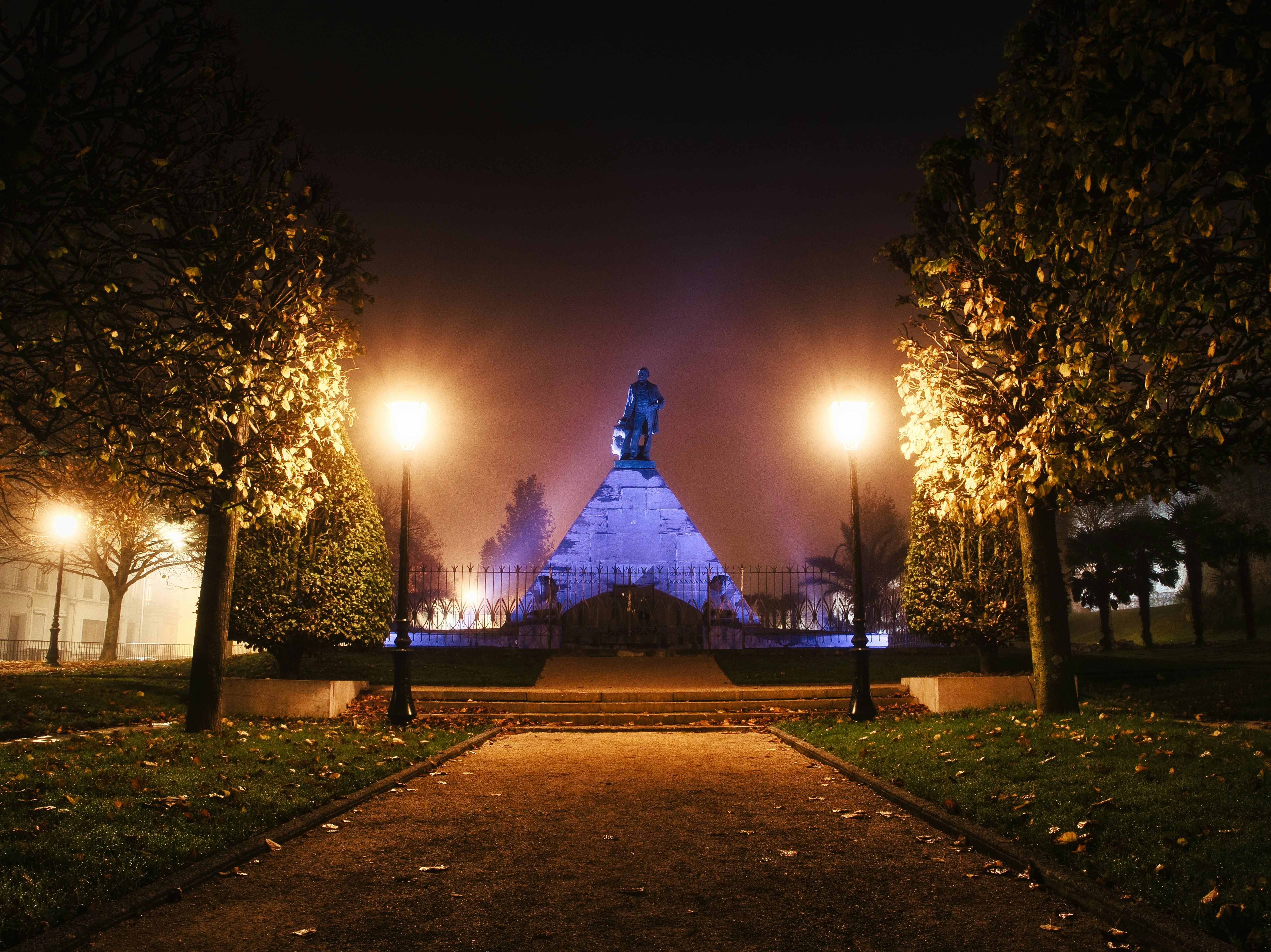 Illuminated monument surrounded by trees and streetlamps in a park at night.