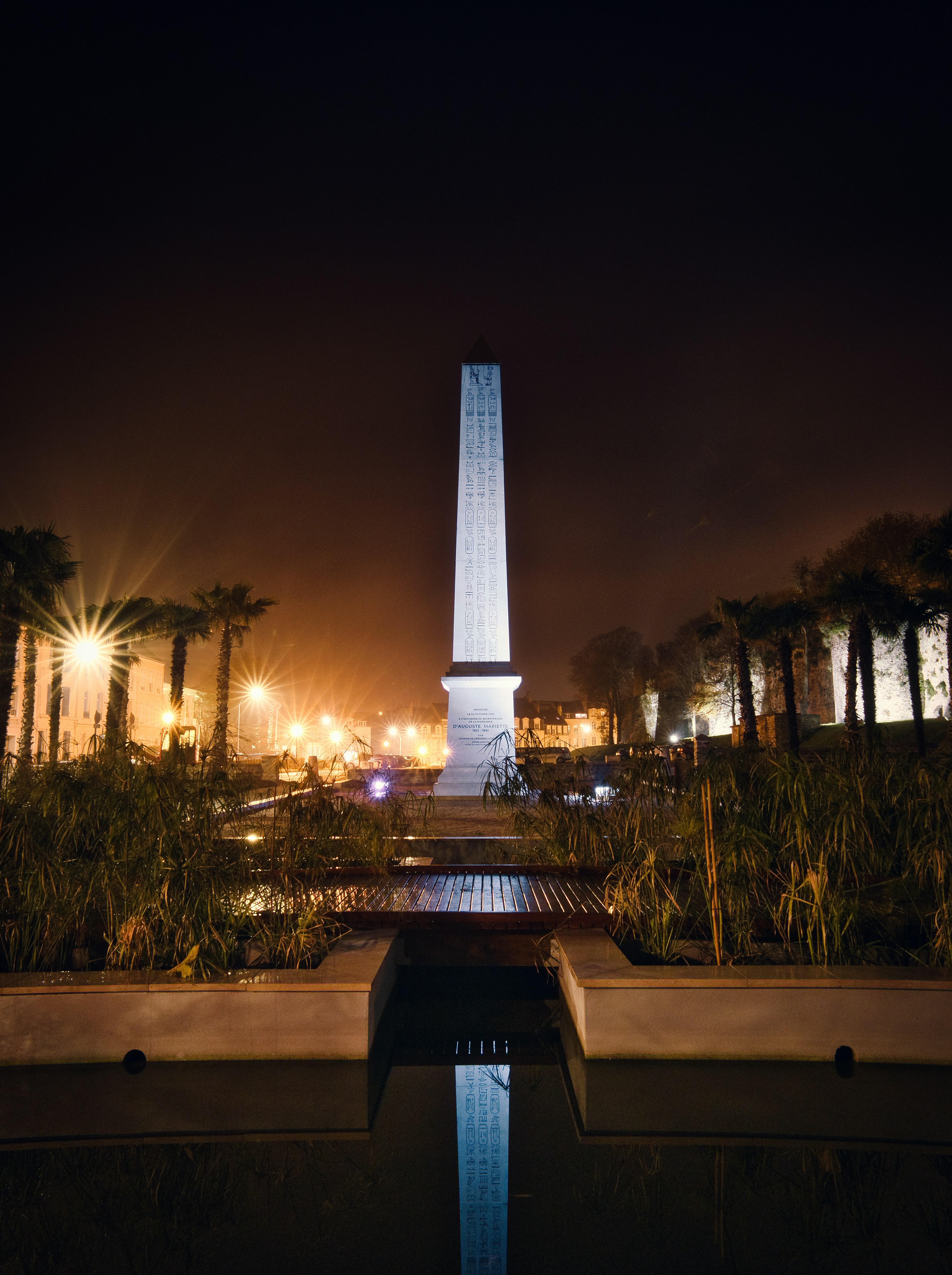 Centered Obelisk Under Warm Night Light: Palm-Lined Plaza and Reflections
