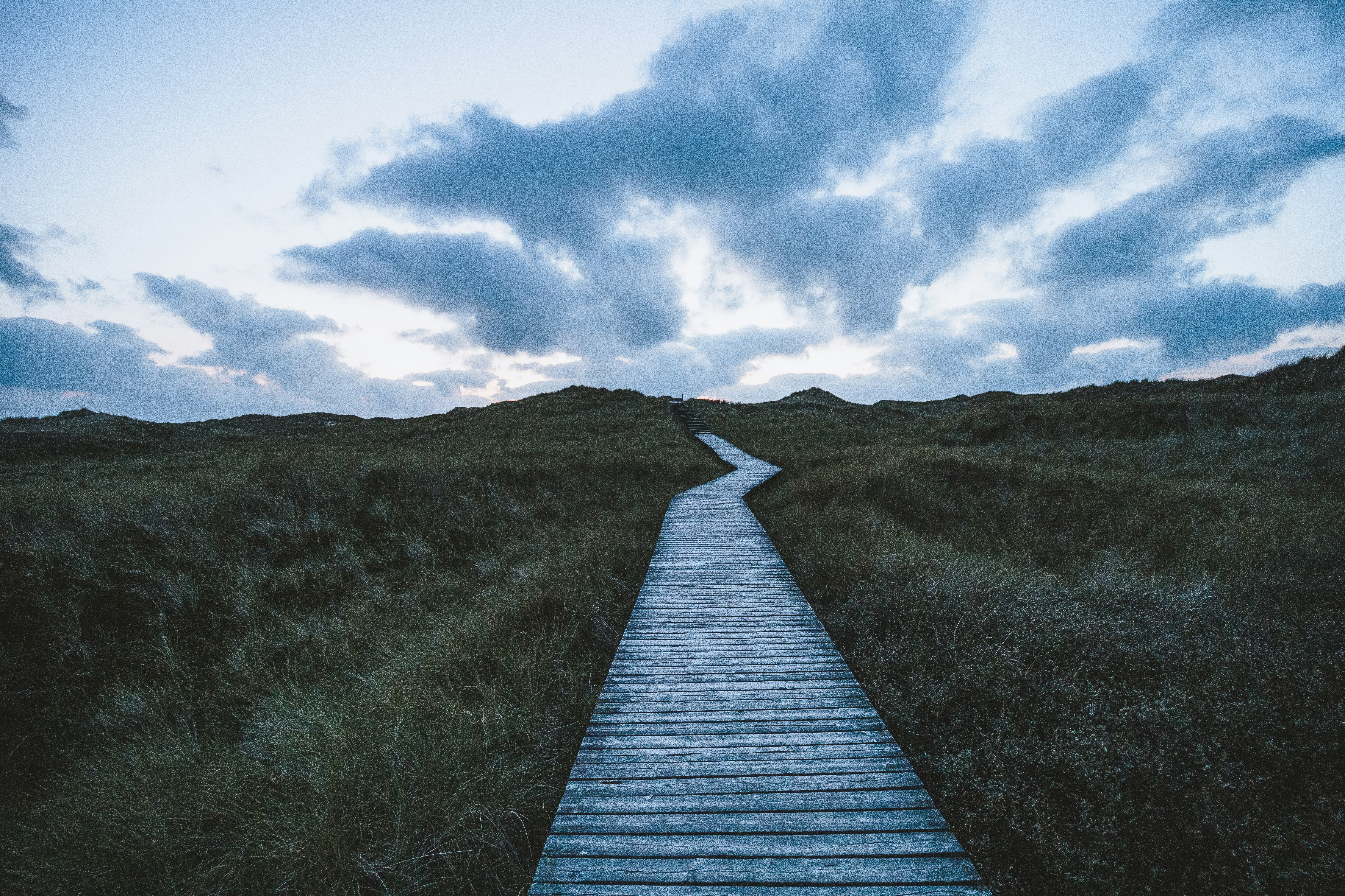 Wooden boardwalk winding through grassy terrain under a cloudy sky at dusk.