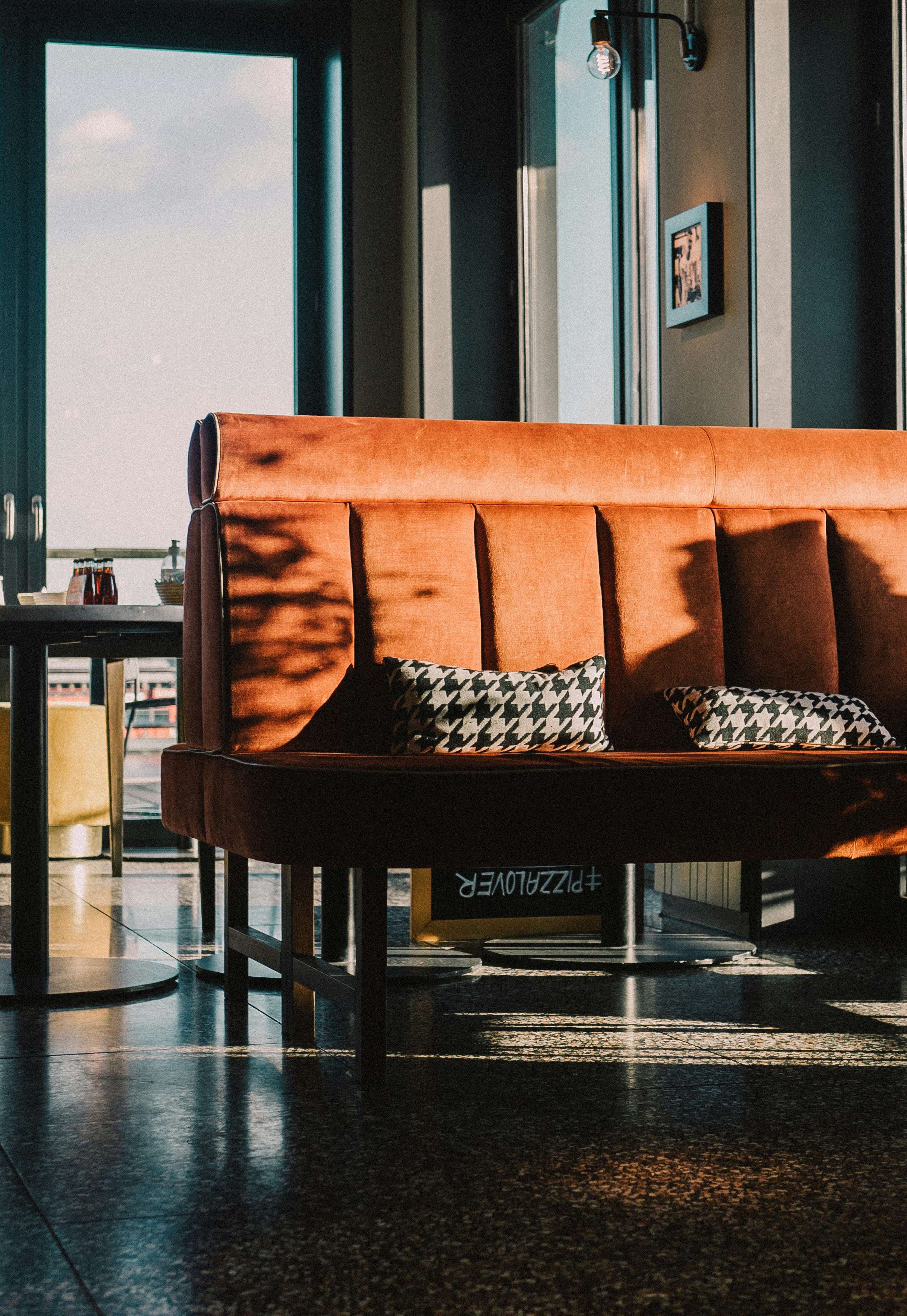 Cozy orange sofa with patterned cushions in a sunlit café, casting shadows on the floor. A refreshing blend of design and ambiance.