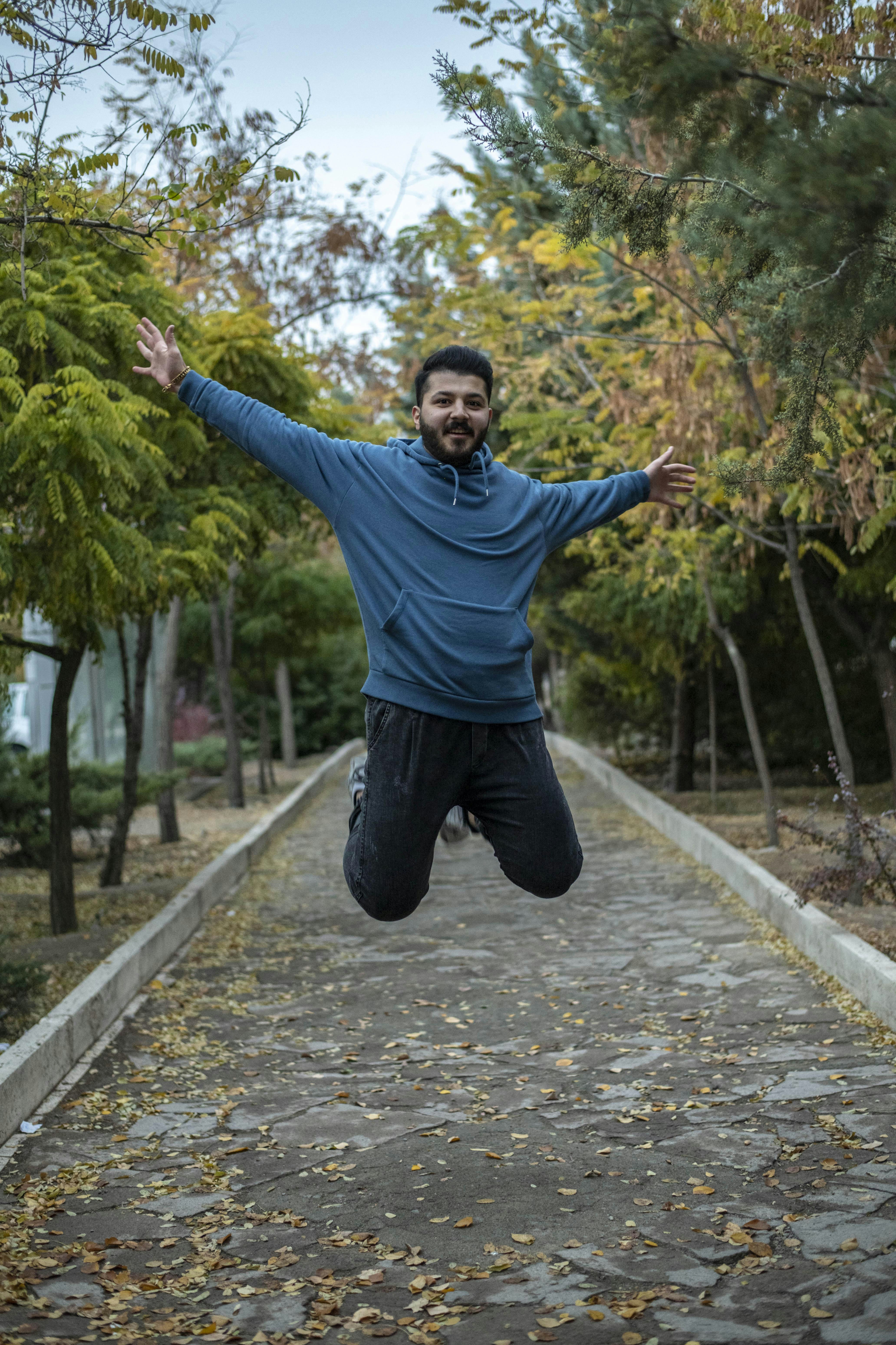 Person joyfully jumping on a stone pathway surrounded by trees with autumn foliage.