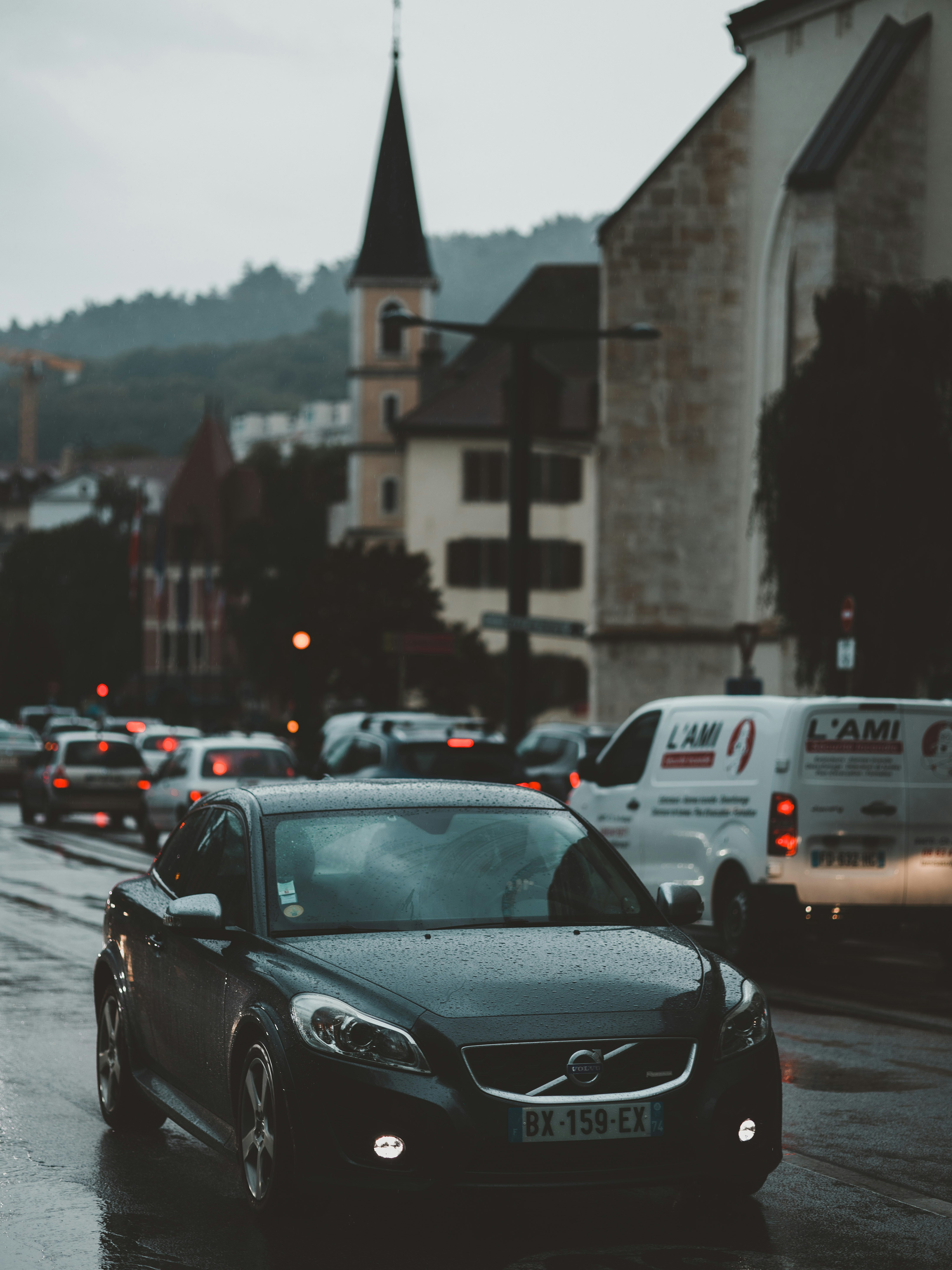 A sleek car navigates through a rain-soaked city street, illuminated by the glow of traffic lights and the silhouette of historic buildings in the background.
