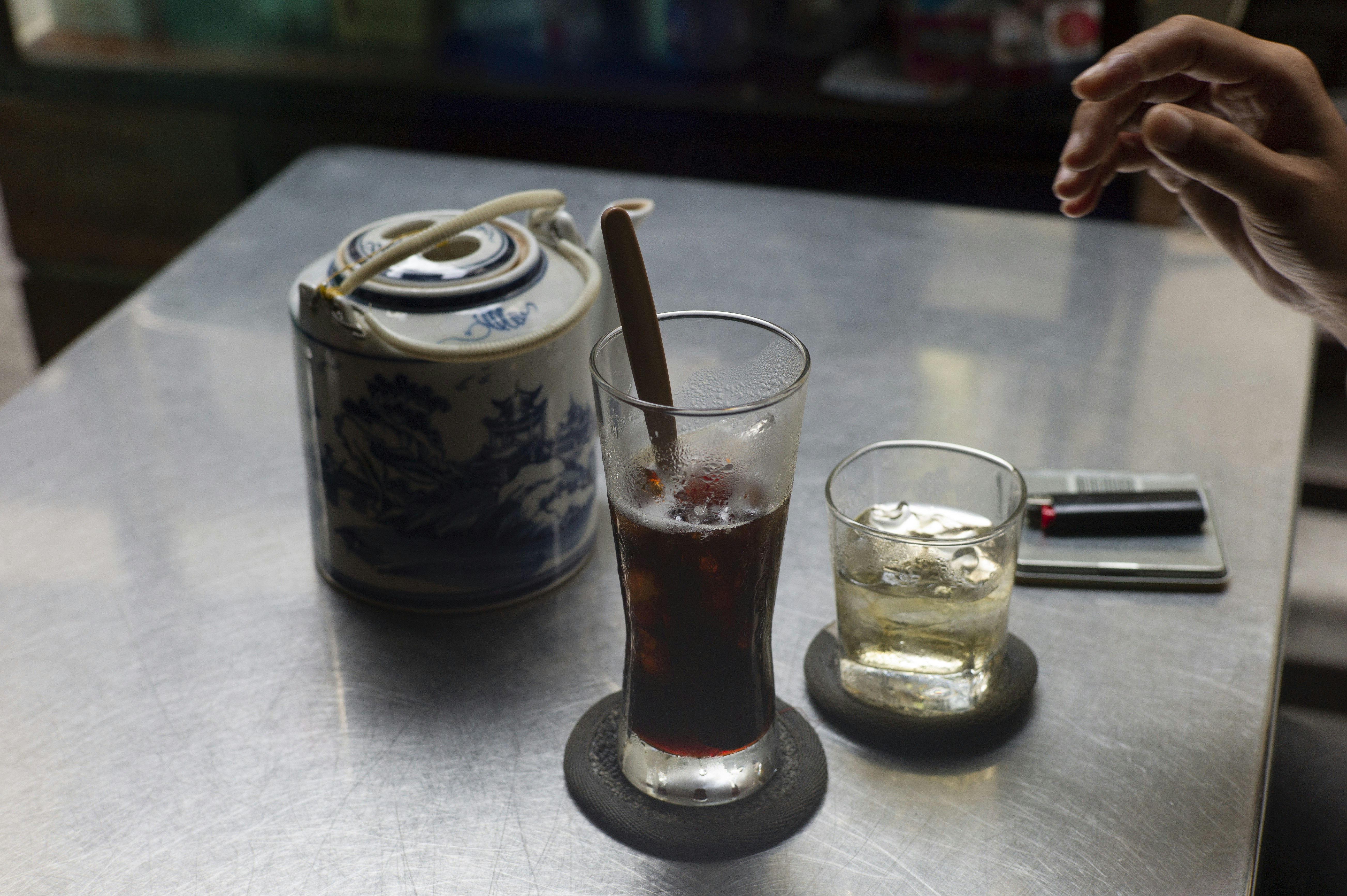 Glass of iced coffee beside a glass of whiskey on a metallic table, with a traditional teapot in the background. A hand gestures nearby.