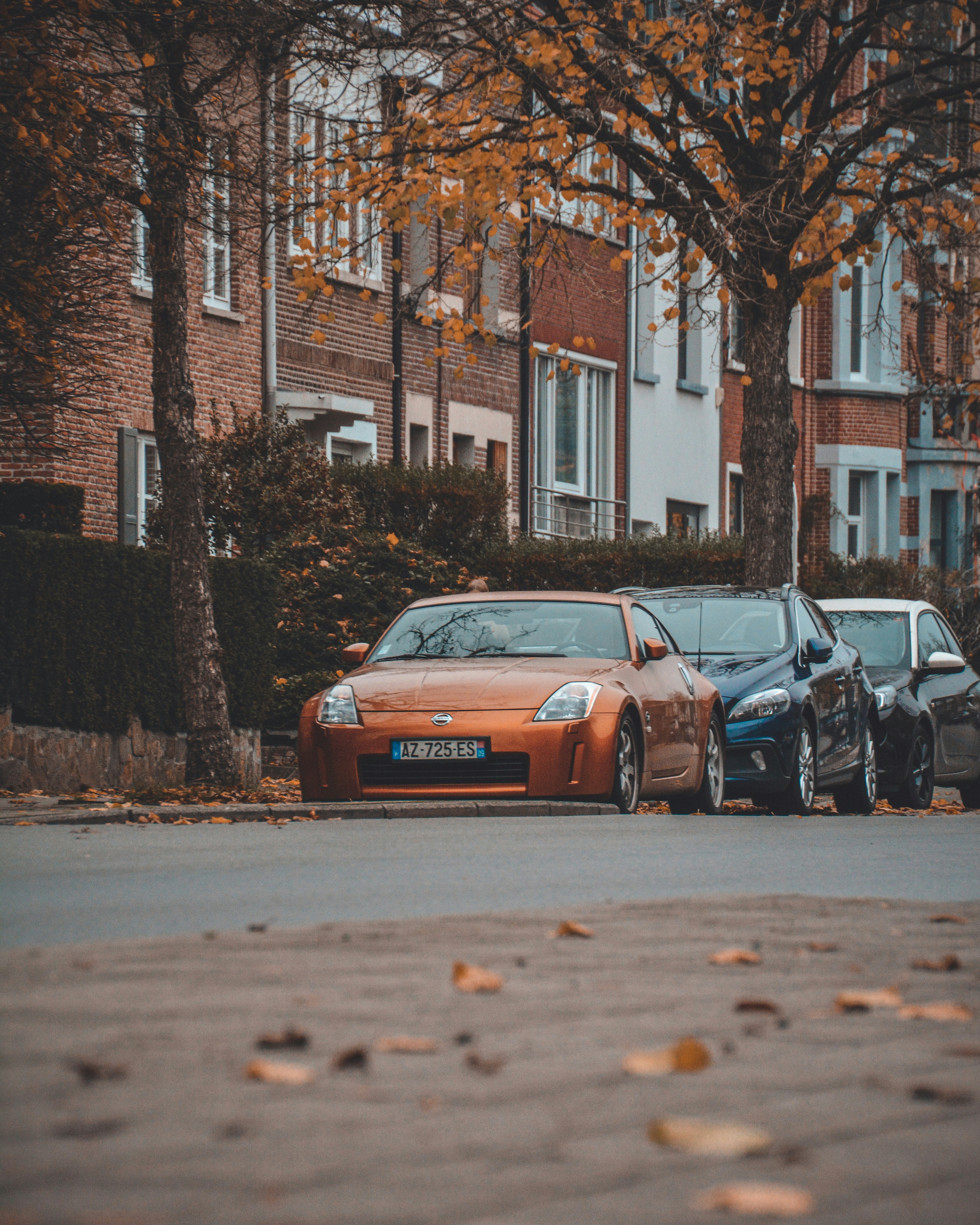 An orange sports car parked on a tree-lined street, surrounded by autumn foliage and residential buildings. Leaves cover the ground, enhancing the seasonal atmosphere.