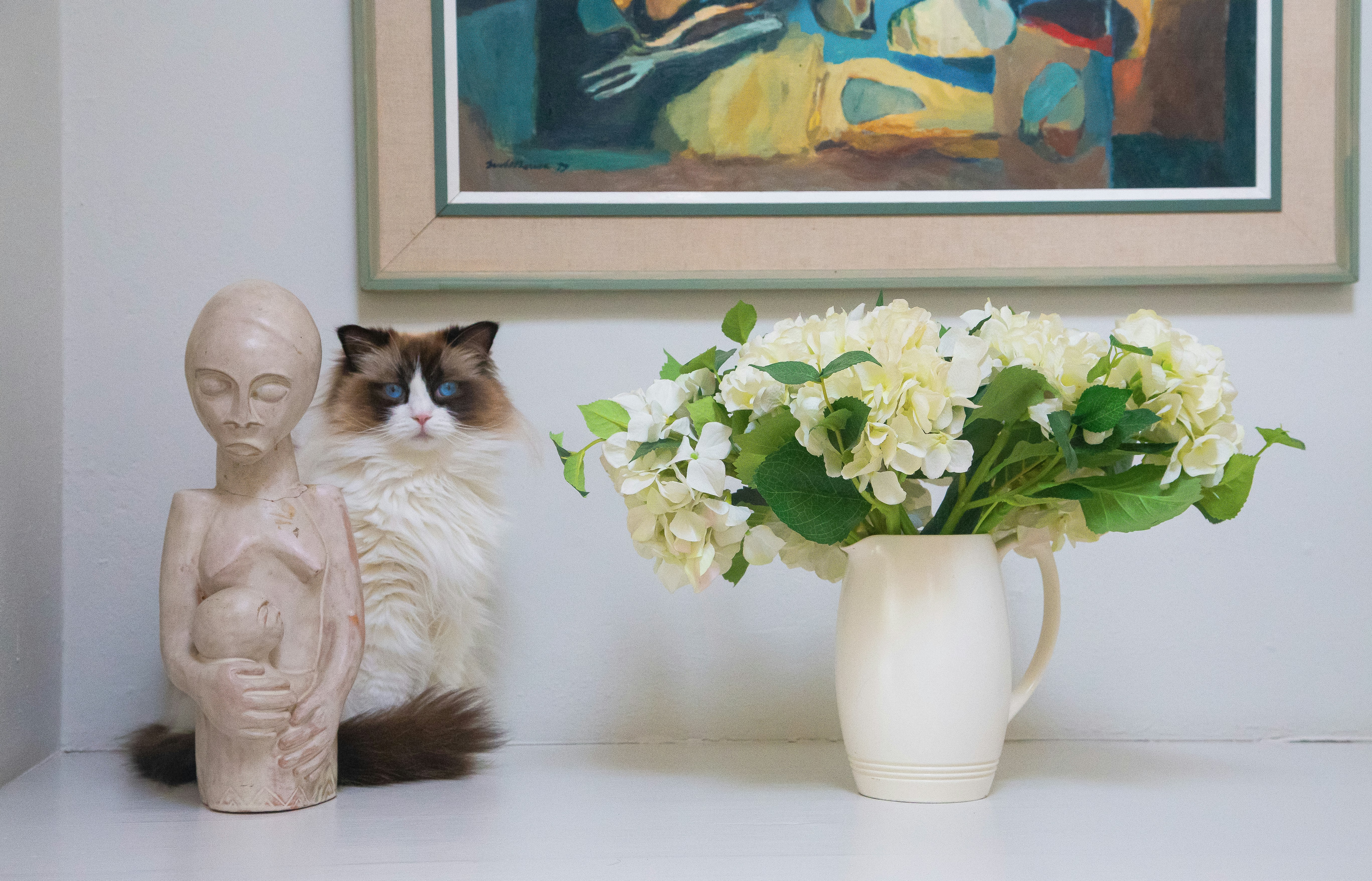Cat nestled between a sculpted figure and a vase of flowers under a framed painting.