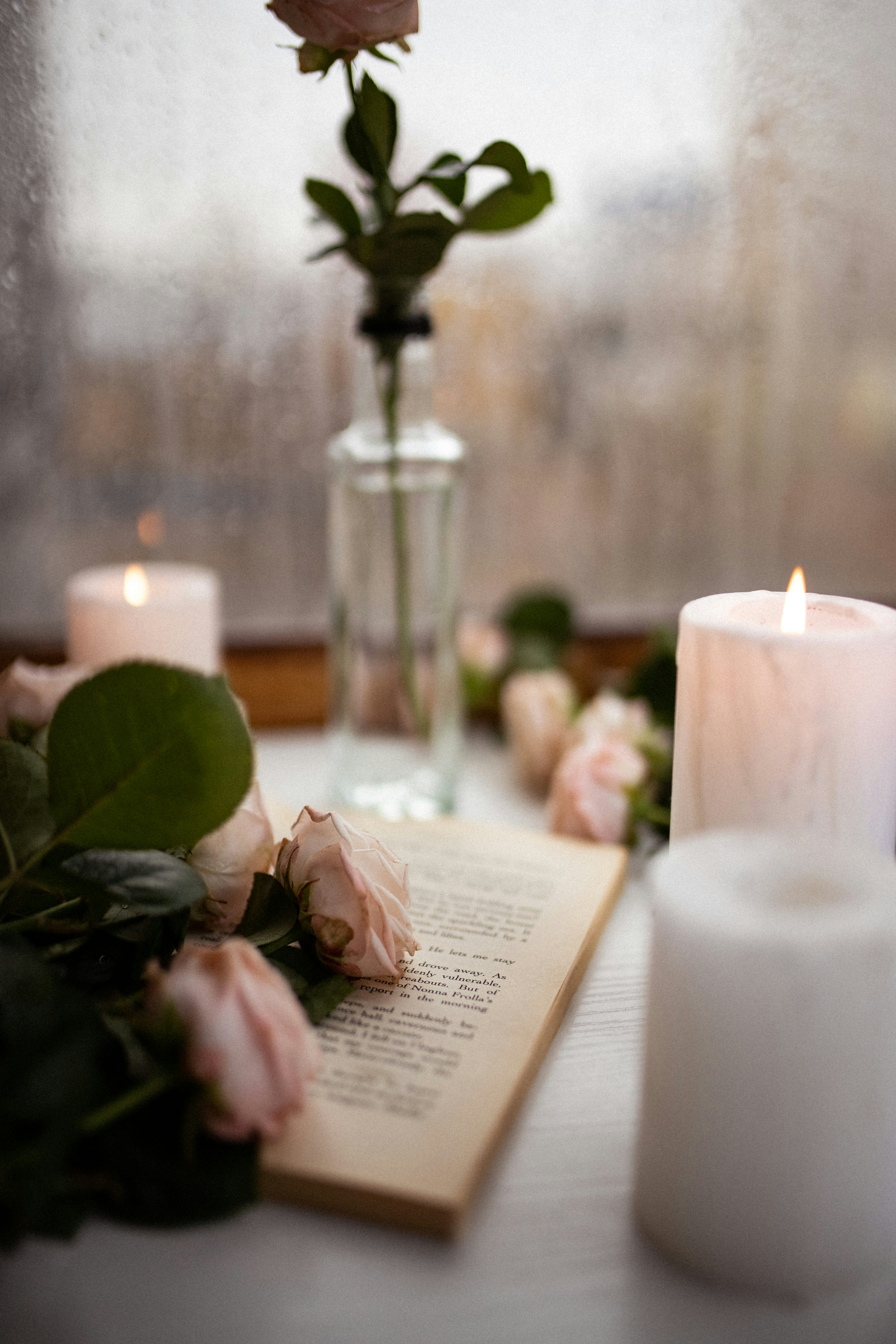 Open book with roses and lit candles on a table by a rain-speckled window.