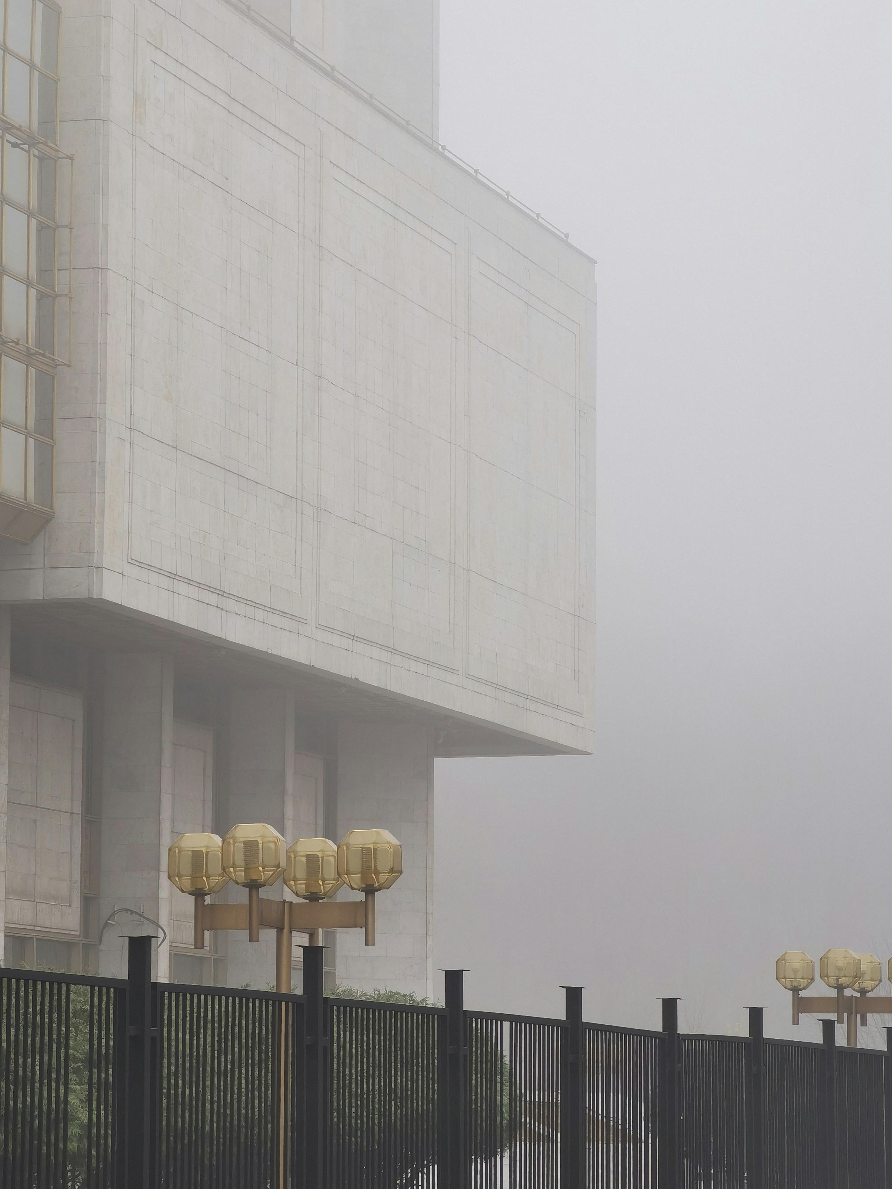 Foggy exterior of a brutalist concrete building with a black metal fence and gold globe street lamps. The scene emphasizes muted detail and an understated atmosphere.