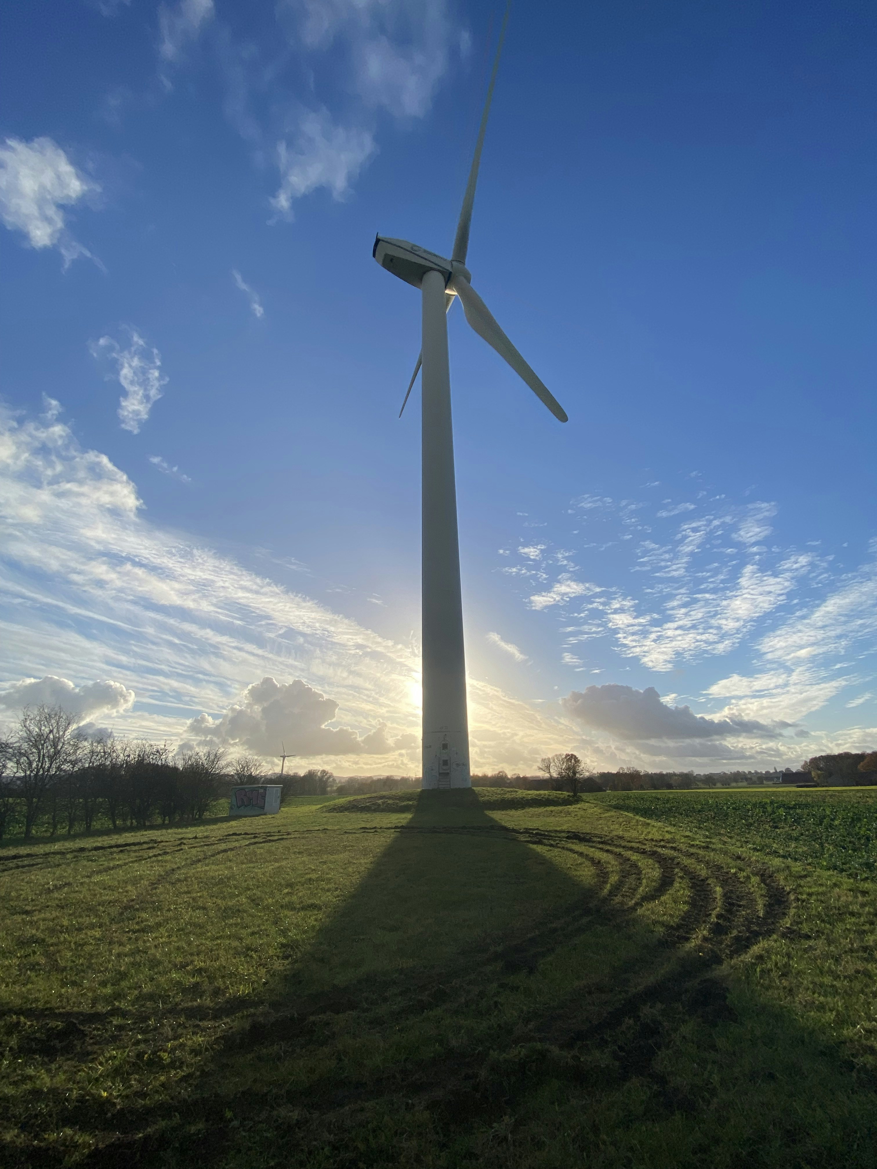 A towering wind turbine stands against a vibrant sky, casting a long shadow over the grassy field. The interplay of light and clouds creates a dynamic backdrop.