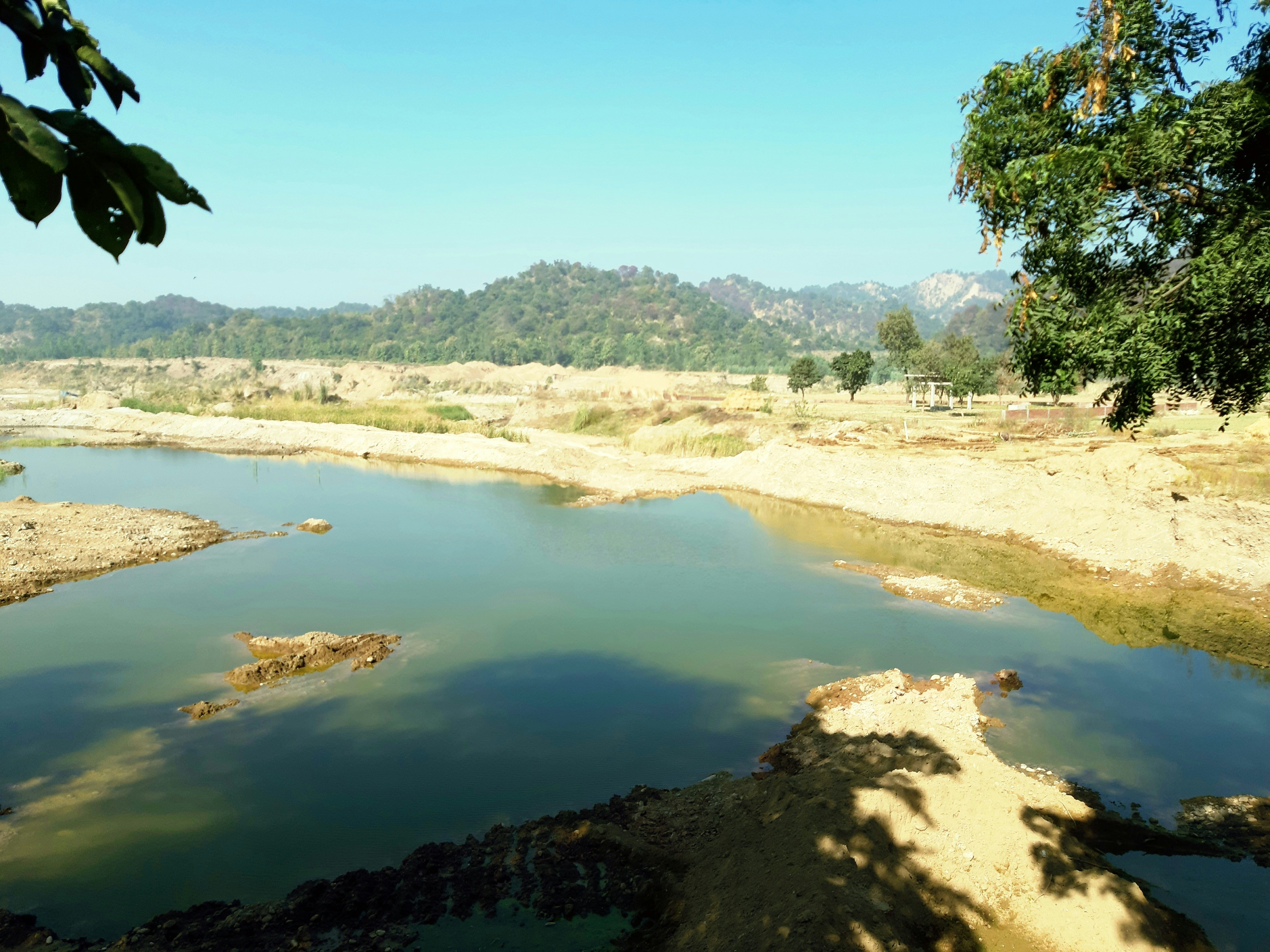 Sunlit river winds through a semi-arid valley, reflecting the clear blue sky. Distant hills form a rolling backdrop to the tranquil water.