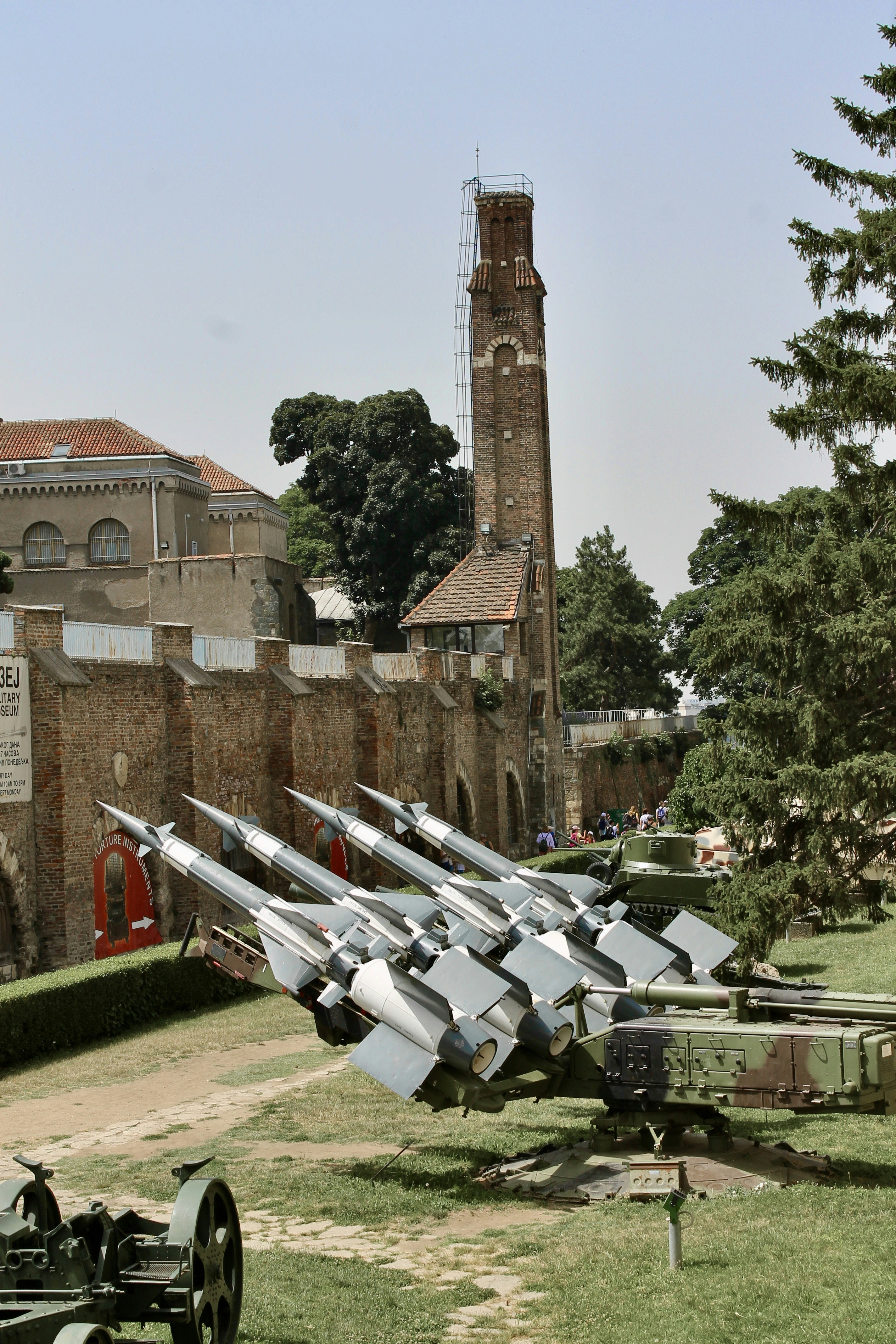 Military artillery pieces arranged in a historical site, showcasing a blend of technology and heritage. The backdrop features an old tower and lush greenery.