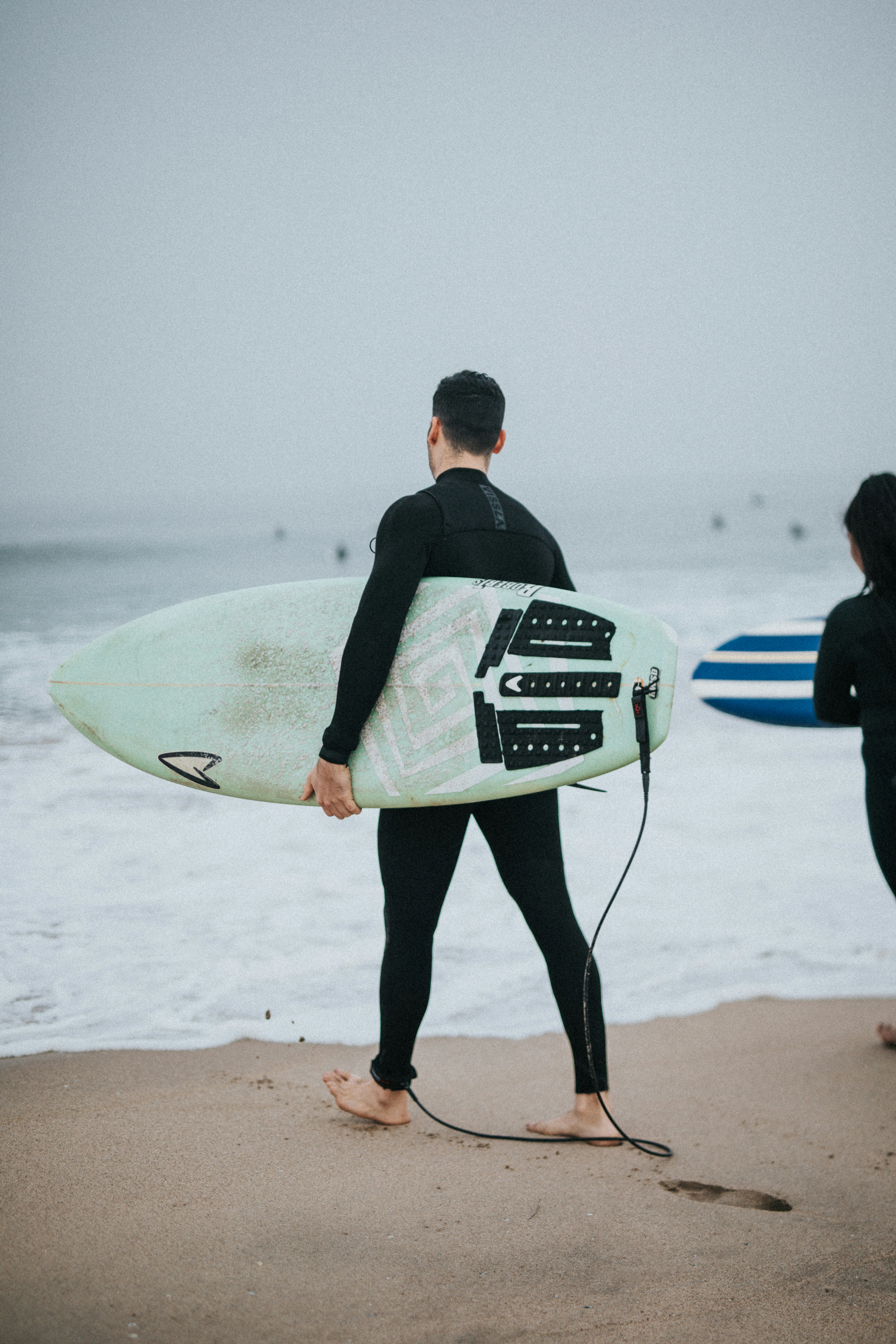 Surfer in a wetsuit holding a green surfboard stands on the beach, gazing at the waves, with another surfer nearby. The scene captures the anticipation of riding the surf.