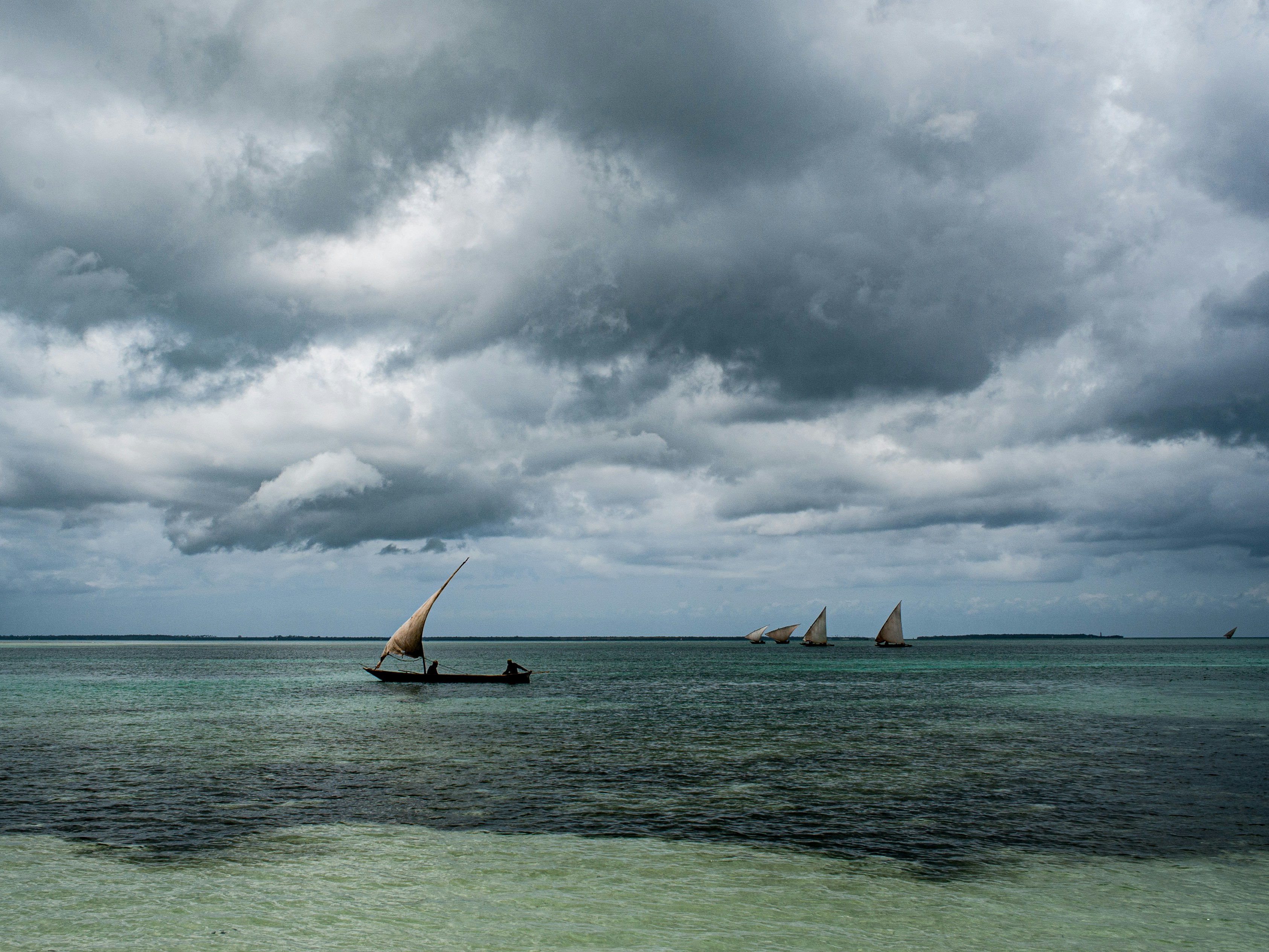 Traditional boats with sails navigate calm waters beneath dramatic, overcast skies.