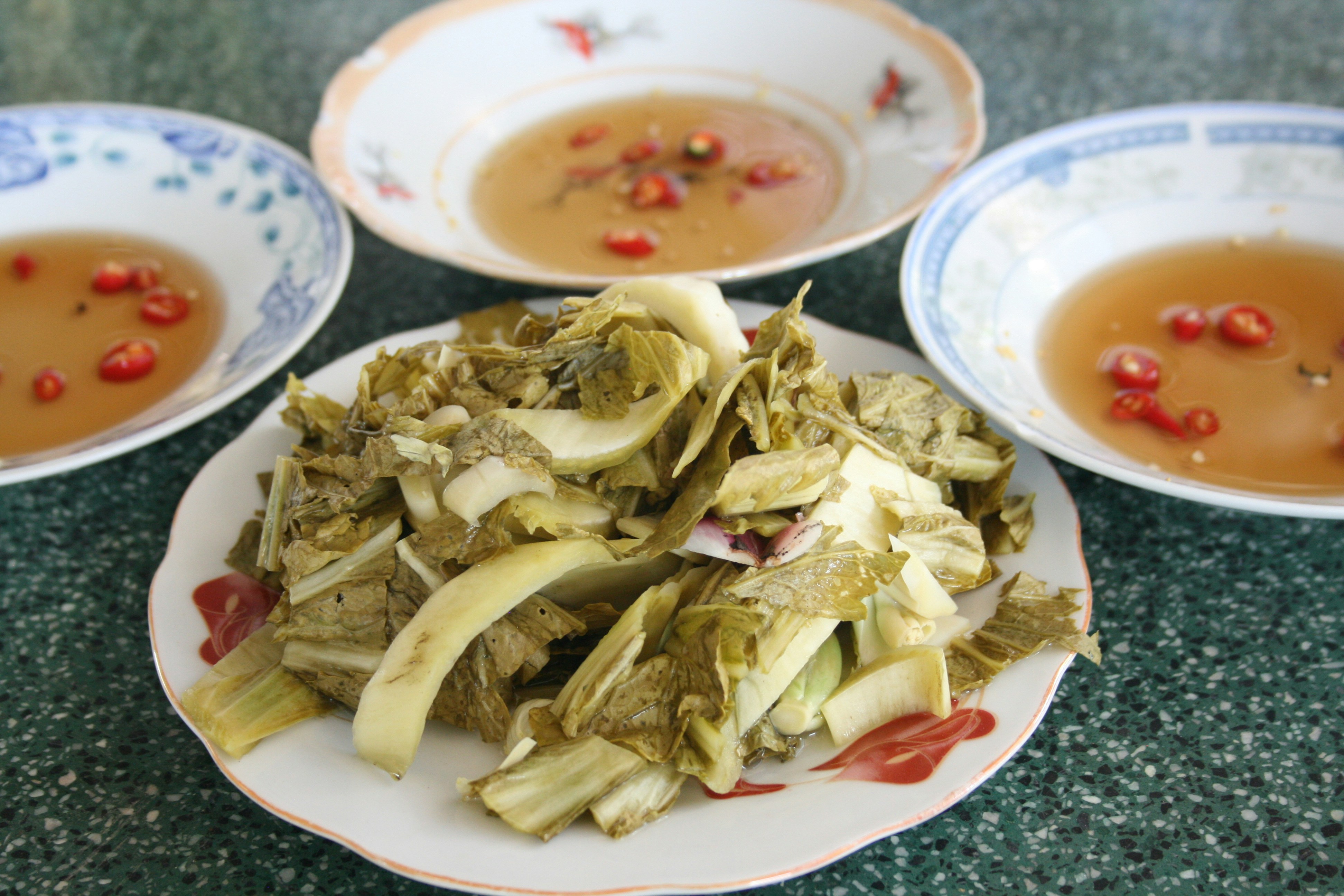 Plate of fresh green vegetables accompanied by three bowls of dipping sauce, each featuring slices of red chili. 