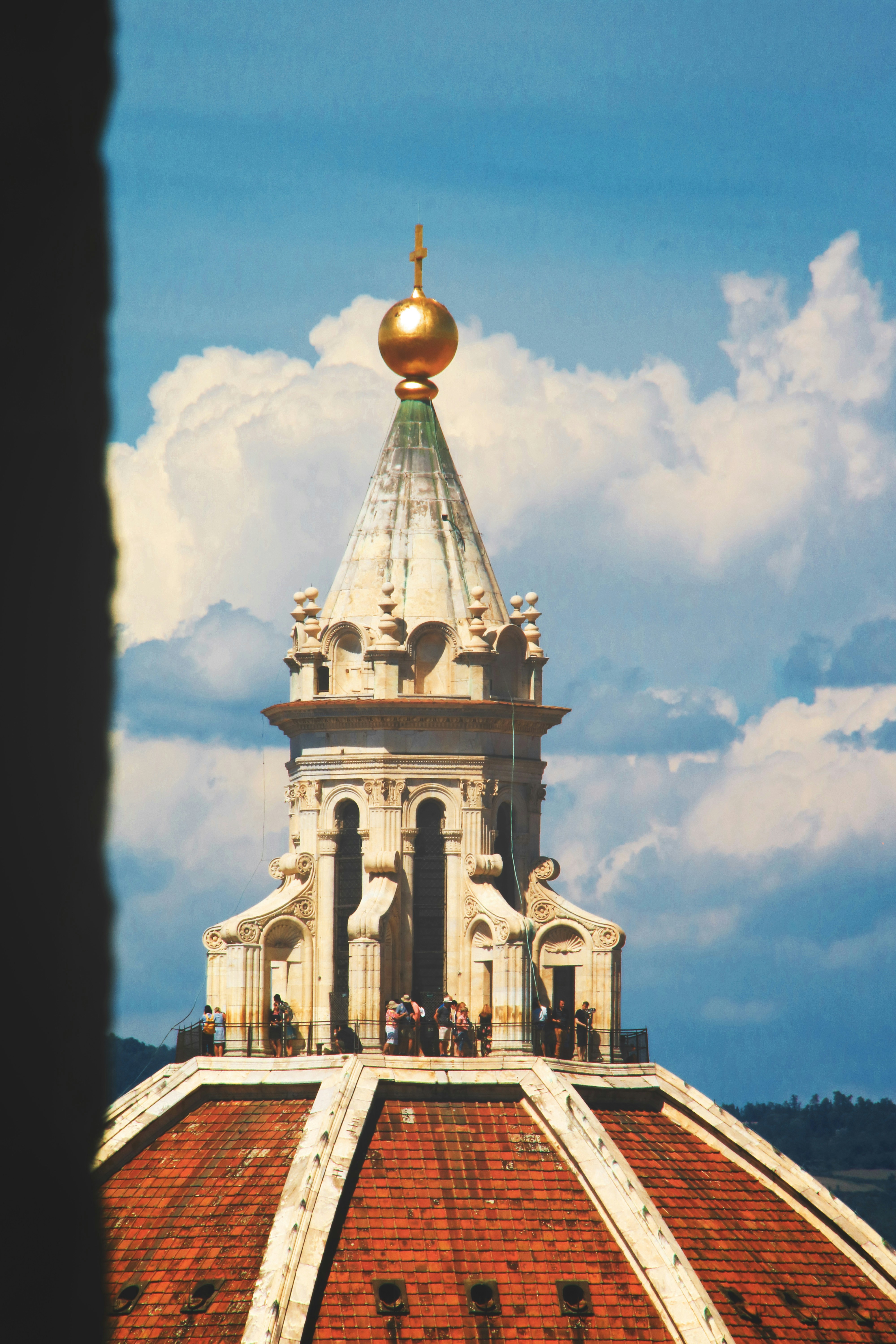 The ornate dome of a historic cathedral, topped with a golden orb, stands against a backdrop of fluffy clouds. Visitors can be seen at the summit, adding a sense of scale.