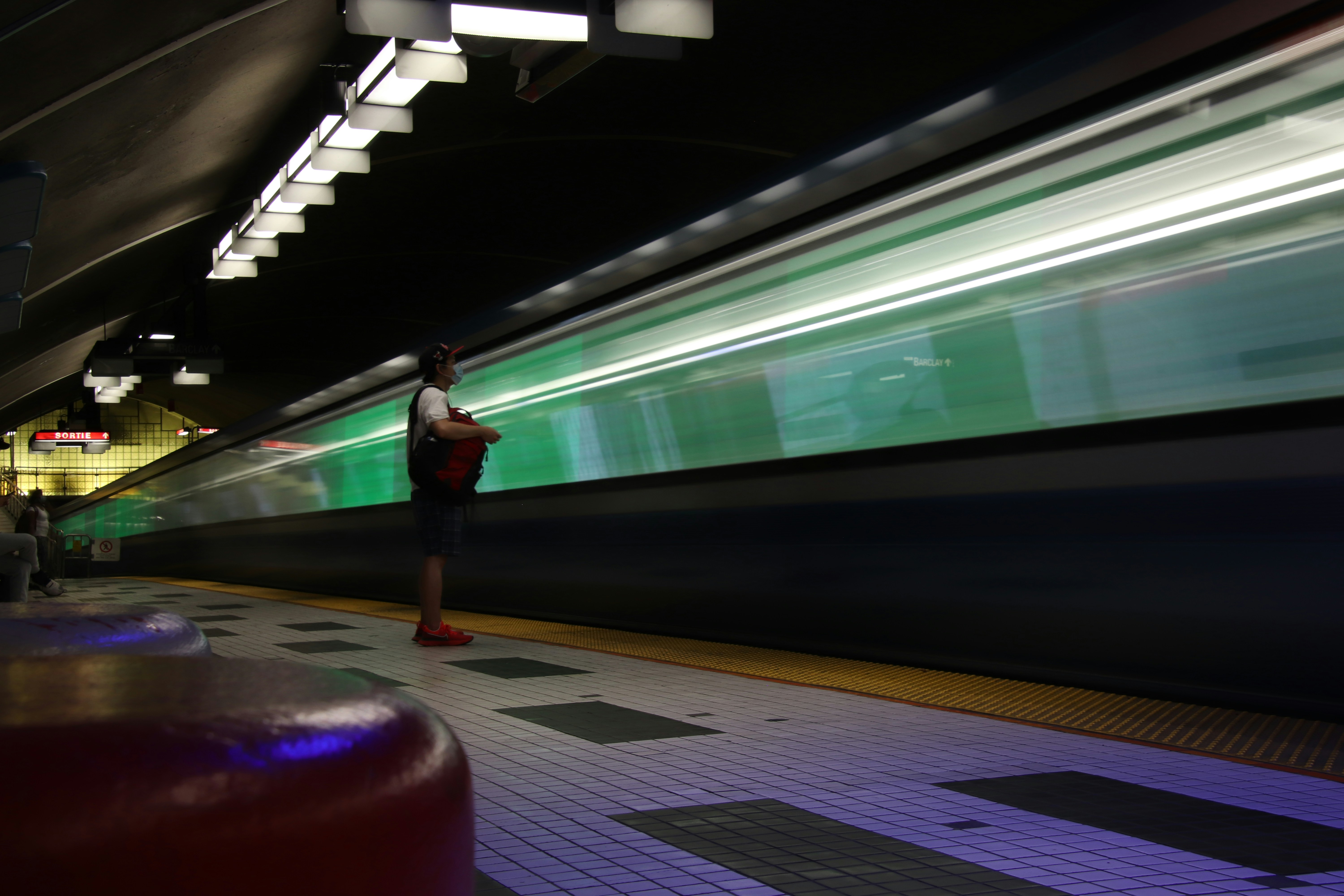 Blurred train rushes past a solitary figure on a dimly lit subway platform.
