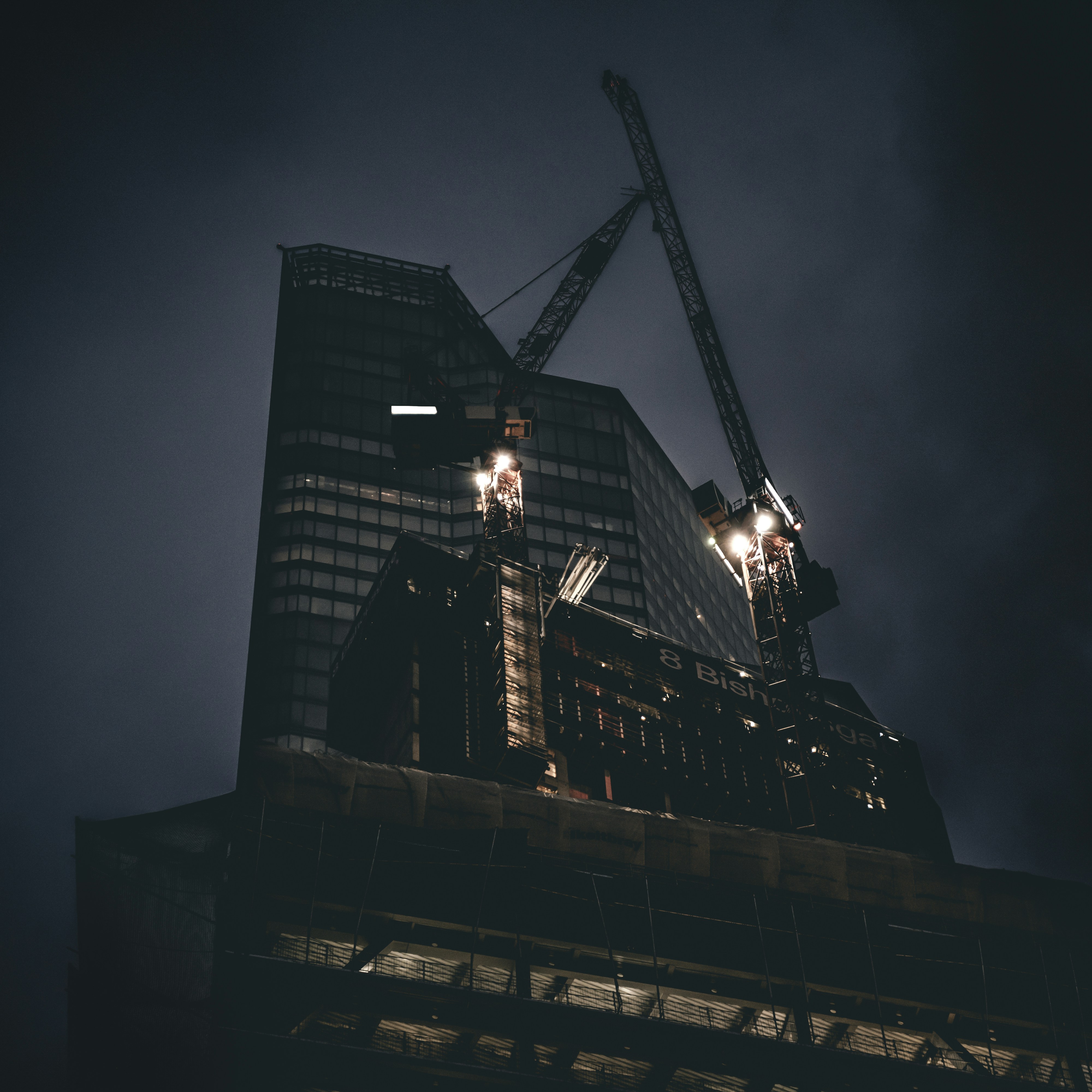 Construction workers maneuvering atop a high-rise building under a moody sky, illuminated by construction lights.