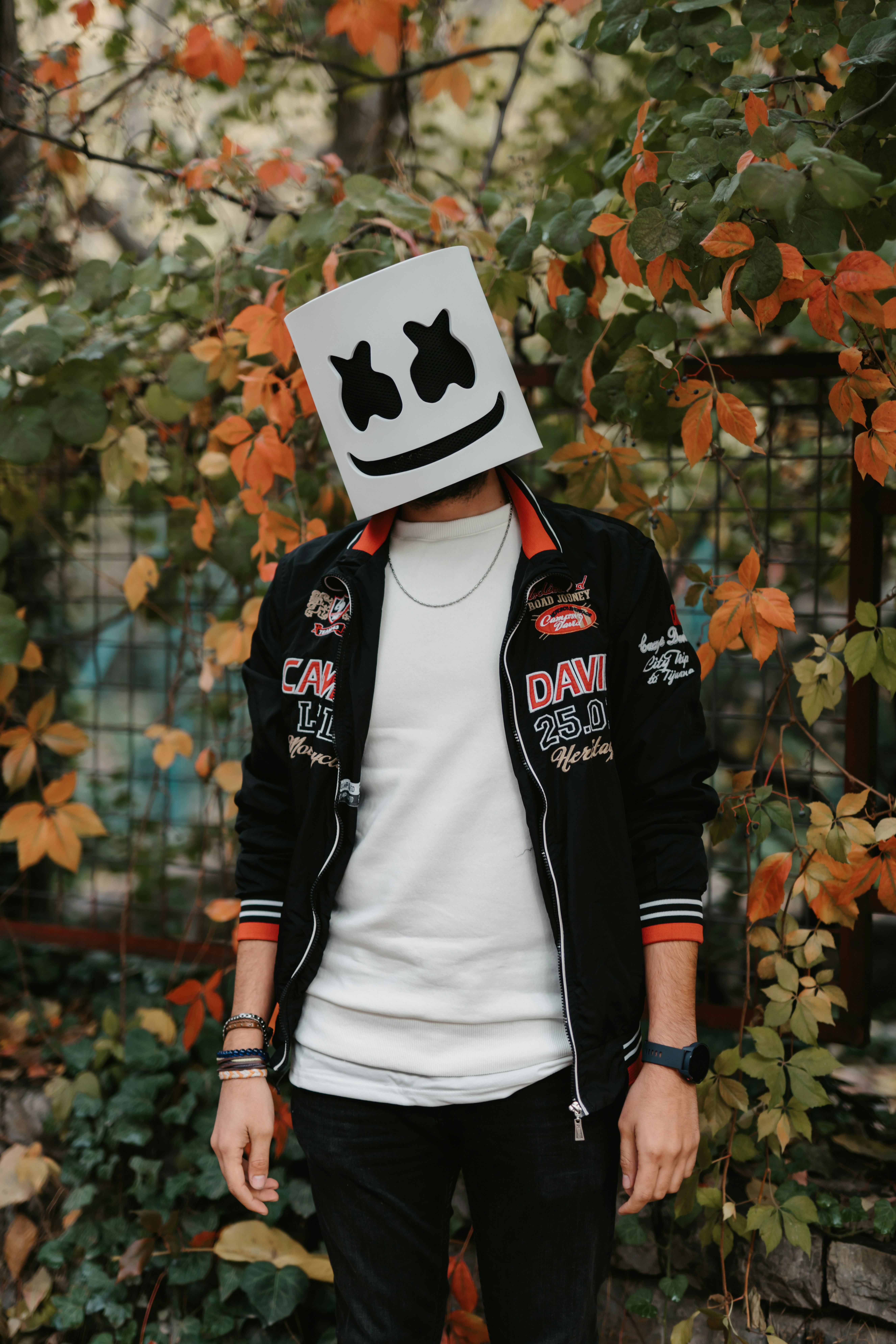 Person with a marshmallow head in a stylish jacket stands among vibrant autumn leaves, creating a playful contrast with the natural backdrop.