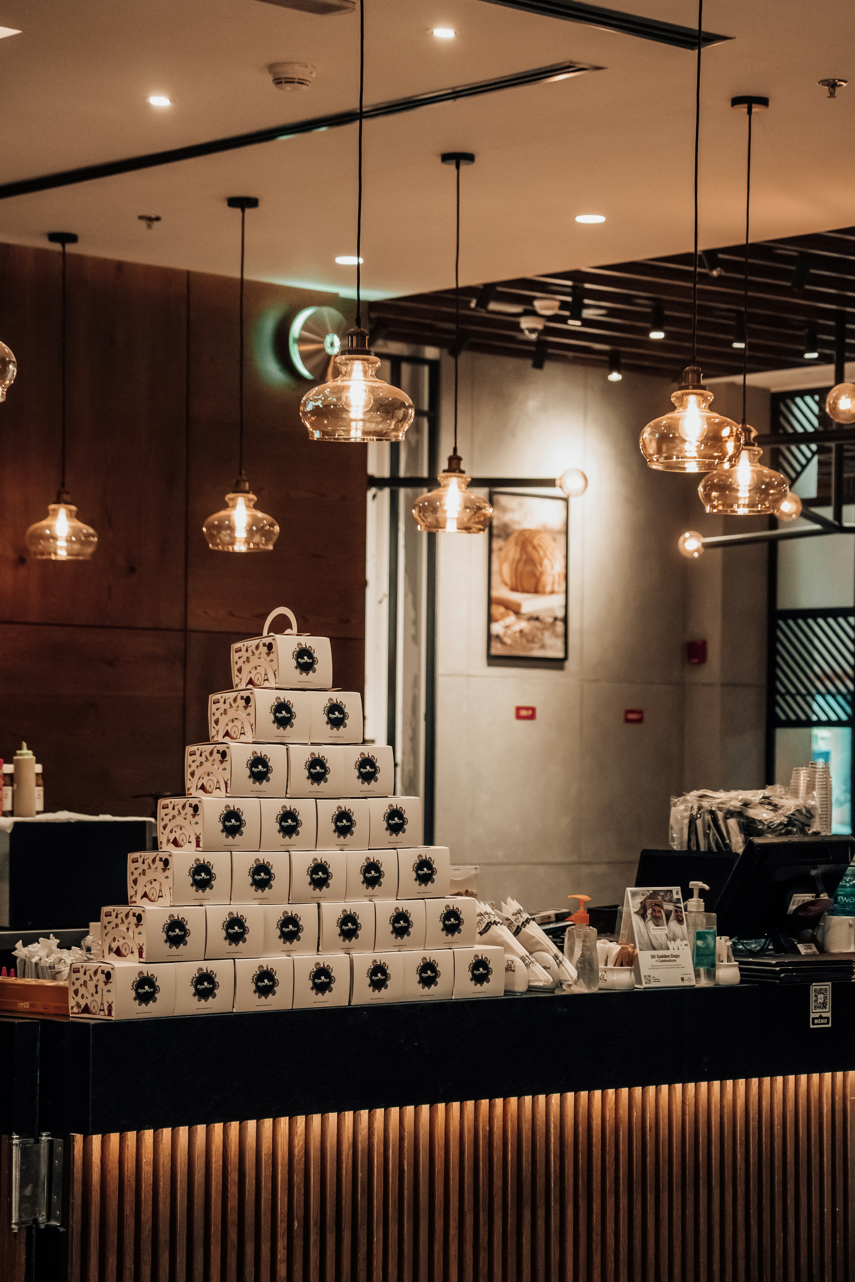 A neatly arranged pyramid of takeout boxes sits prominently on a counter, illuminated by warm pendant lights in a contemporary café setting.