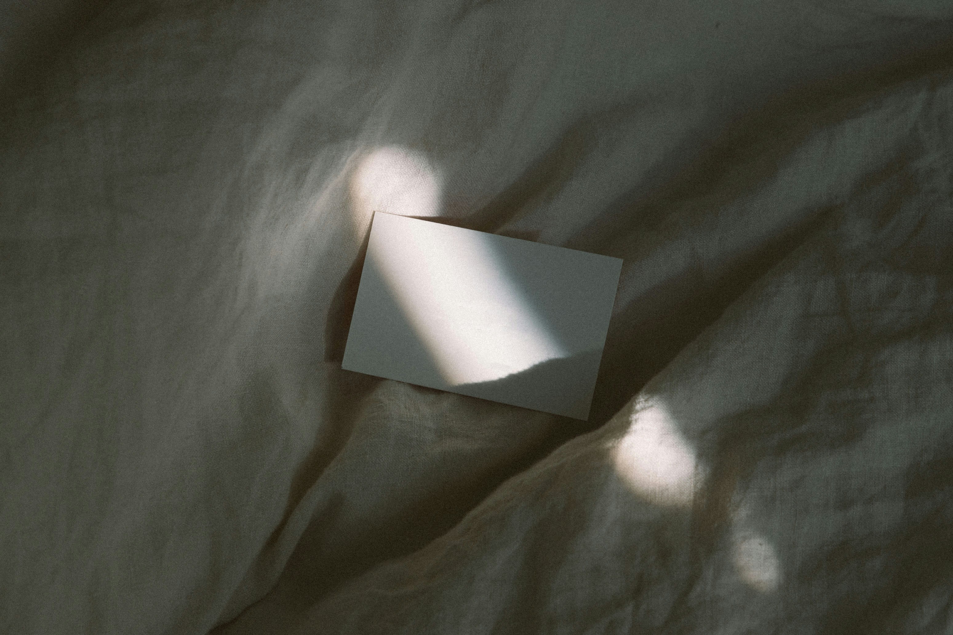 Minimalist composition featuring a blank card resting on soft fabric, illuminated by gentle sunlight creating delicate shadows.