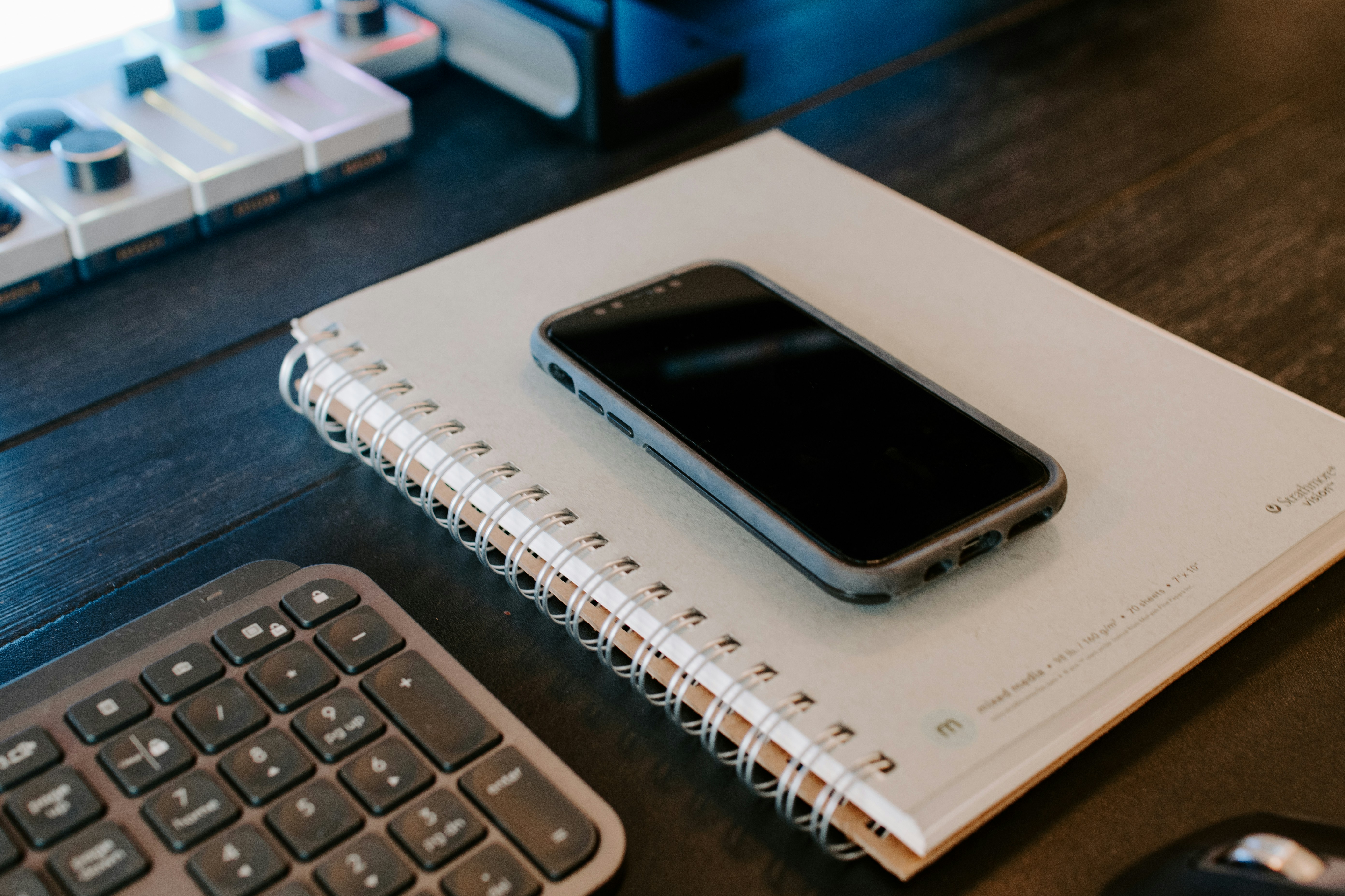 Smartphone resting on a spiral notebook in a contemporary workspace, surrounded by a keyboard and audio equipment.