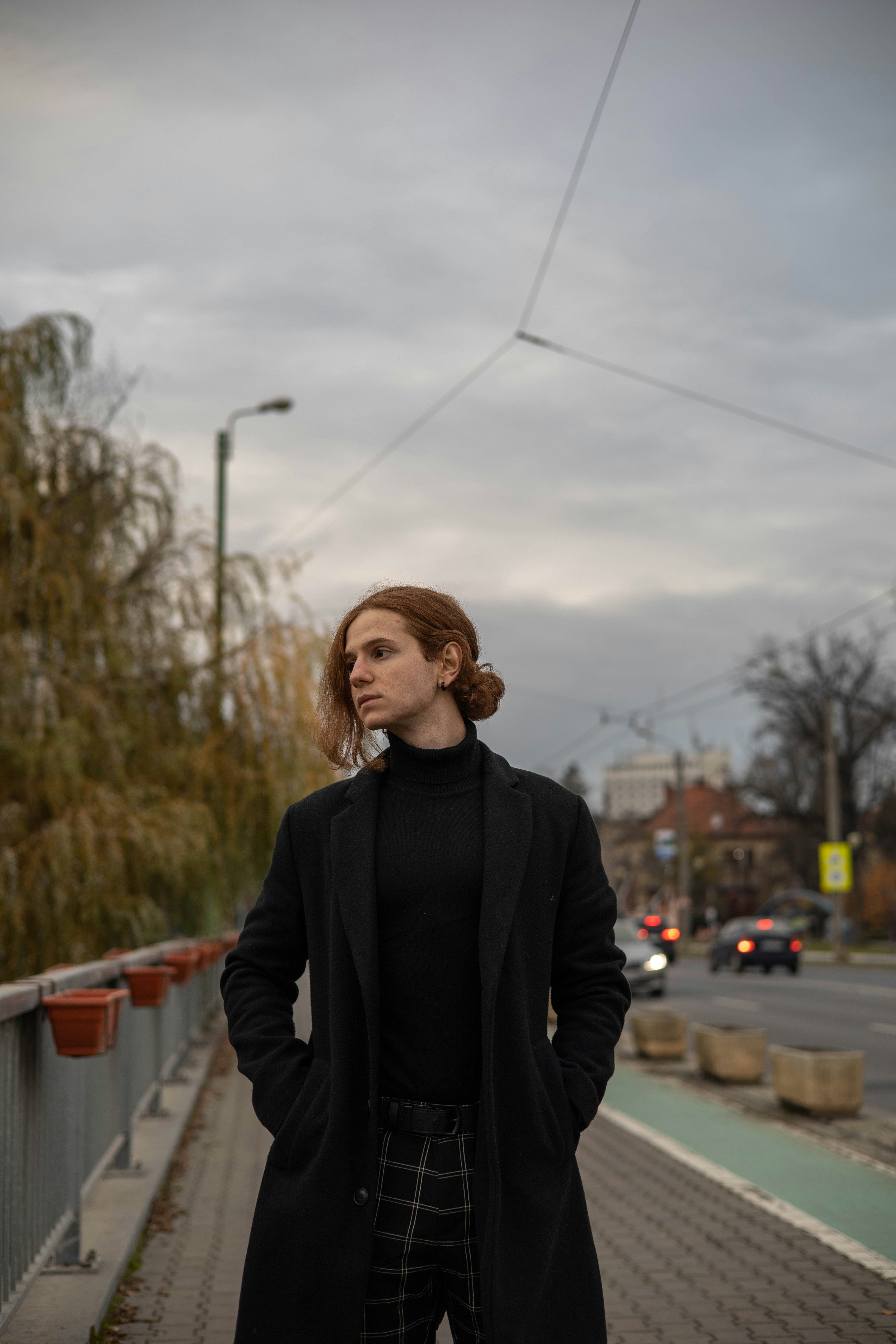 A young man stands thoughtfully on a city sidewalk, dressed in a black coat and turtleneck, surrounded by urban elements and soft evening light.