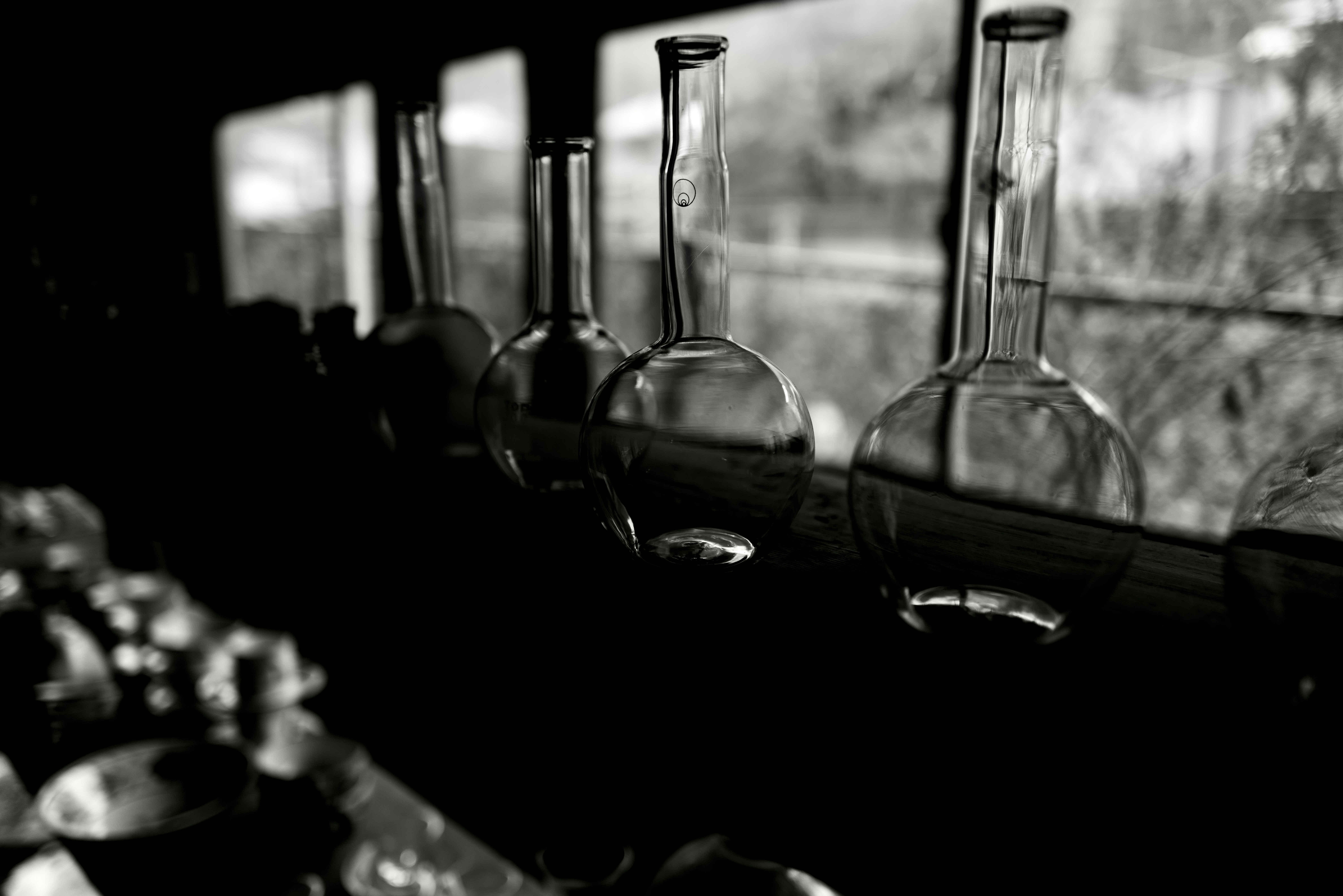 A collection of glass laboratory flasks arranged on a windowsill, capturing the interplay of light and shadows. The monochrome setting enhances the textures and shapes of the glassware.