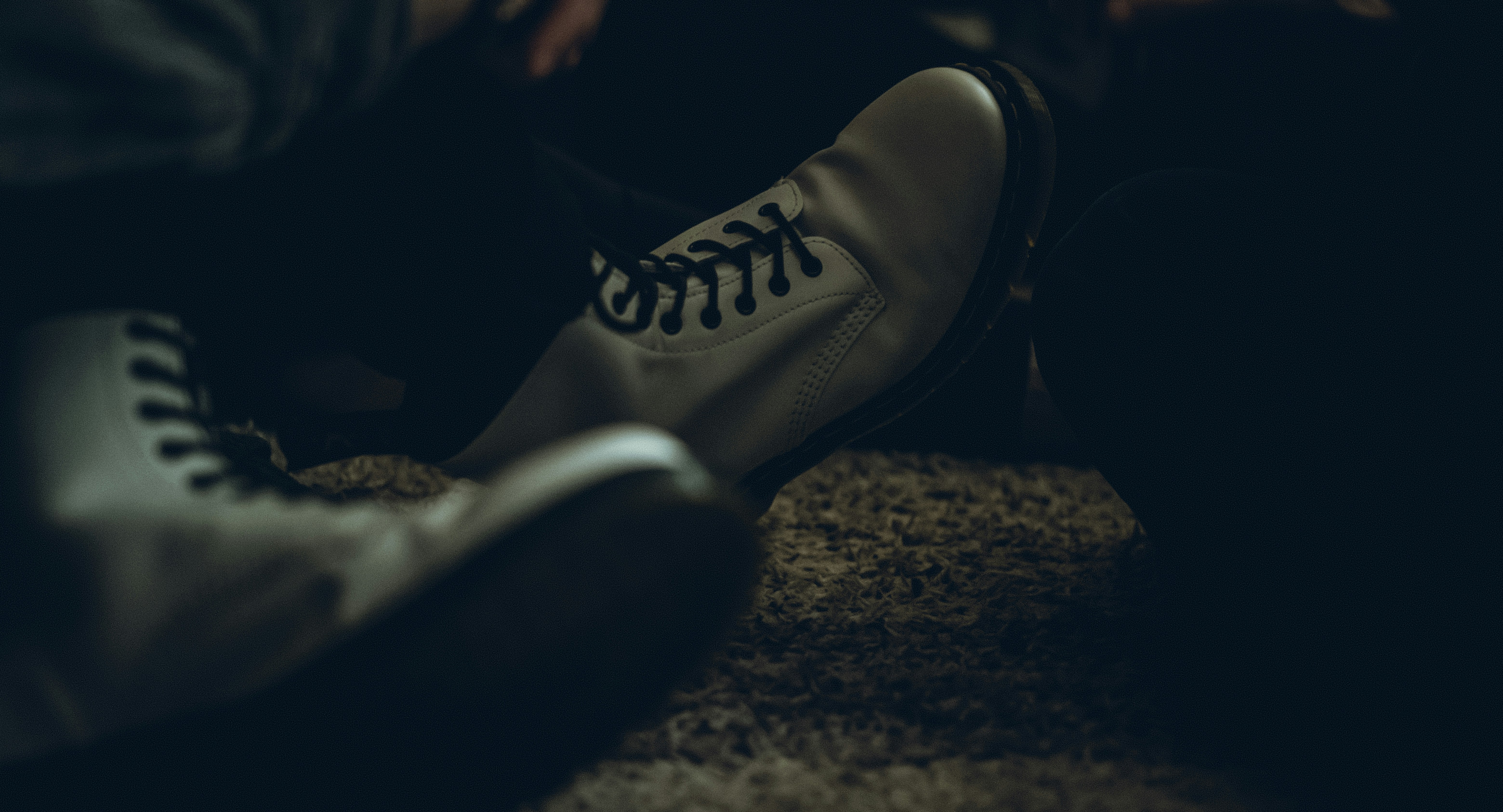Close-up of white lace-up boots resting on a textured surface, surrounded by shadowy figures. The scene conveys an intimate moment of togetherness.