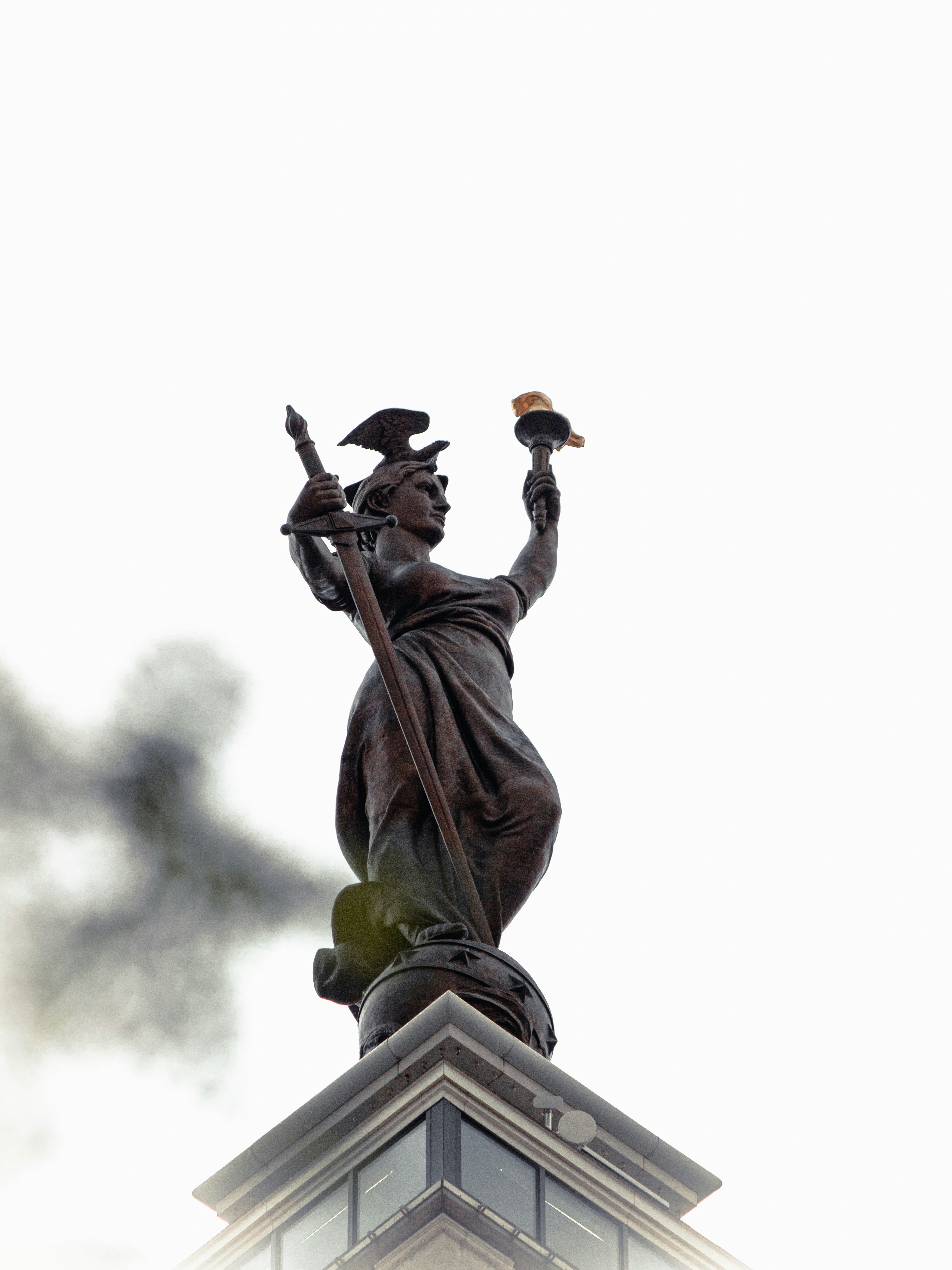 Bronze statue of a robed figure holding a torch and a staff, symbolizing justice, atop a building against a bright sky.