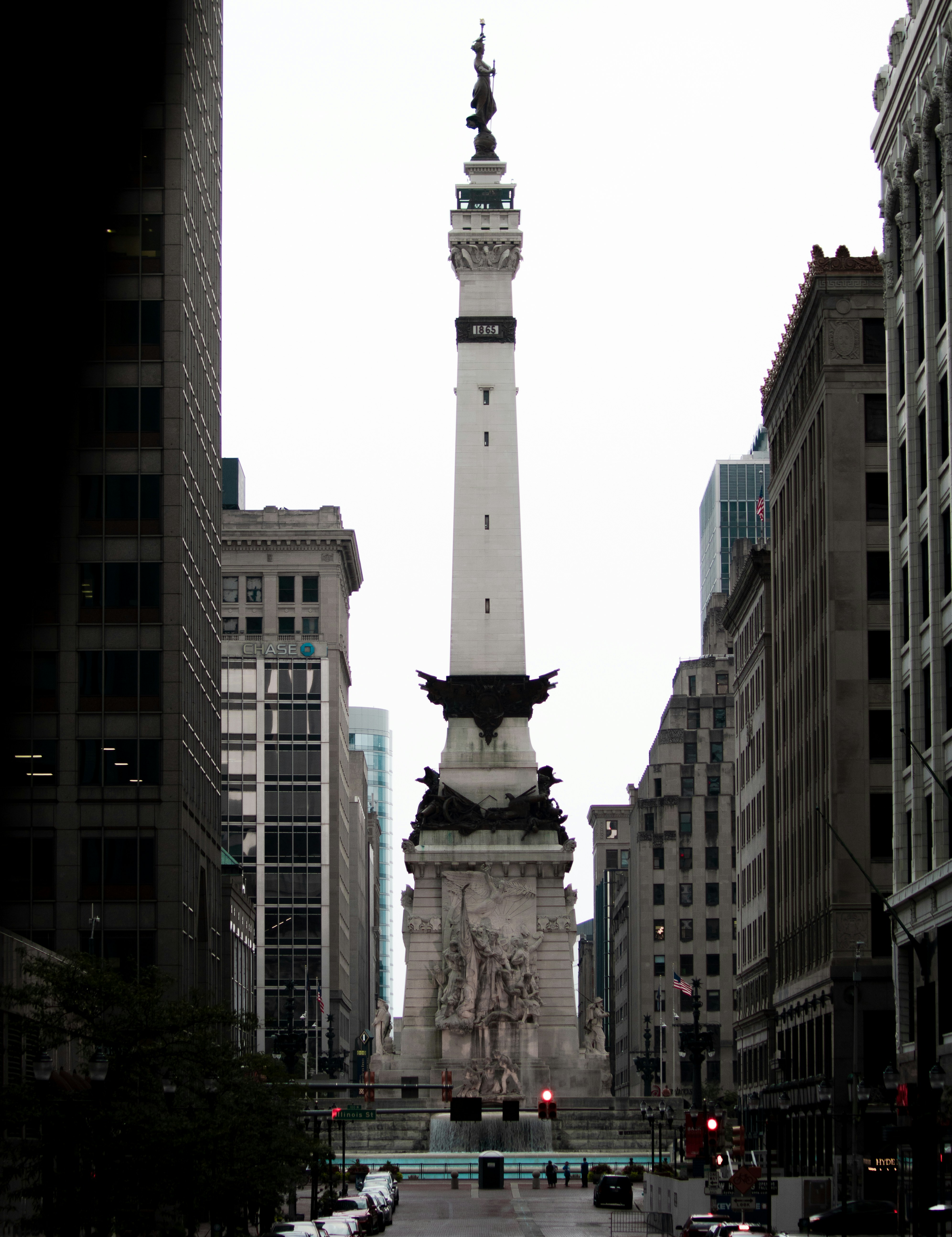 A towering war memorial stands at the end of a city street, flanked by modern skyscrapers and bathed in muted light.